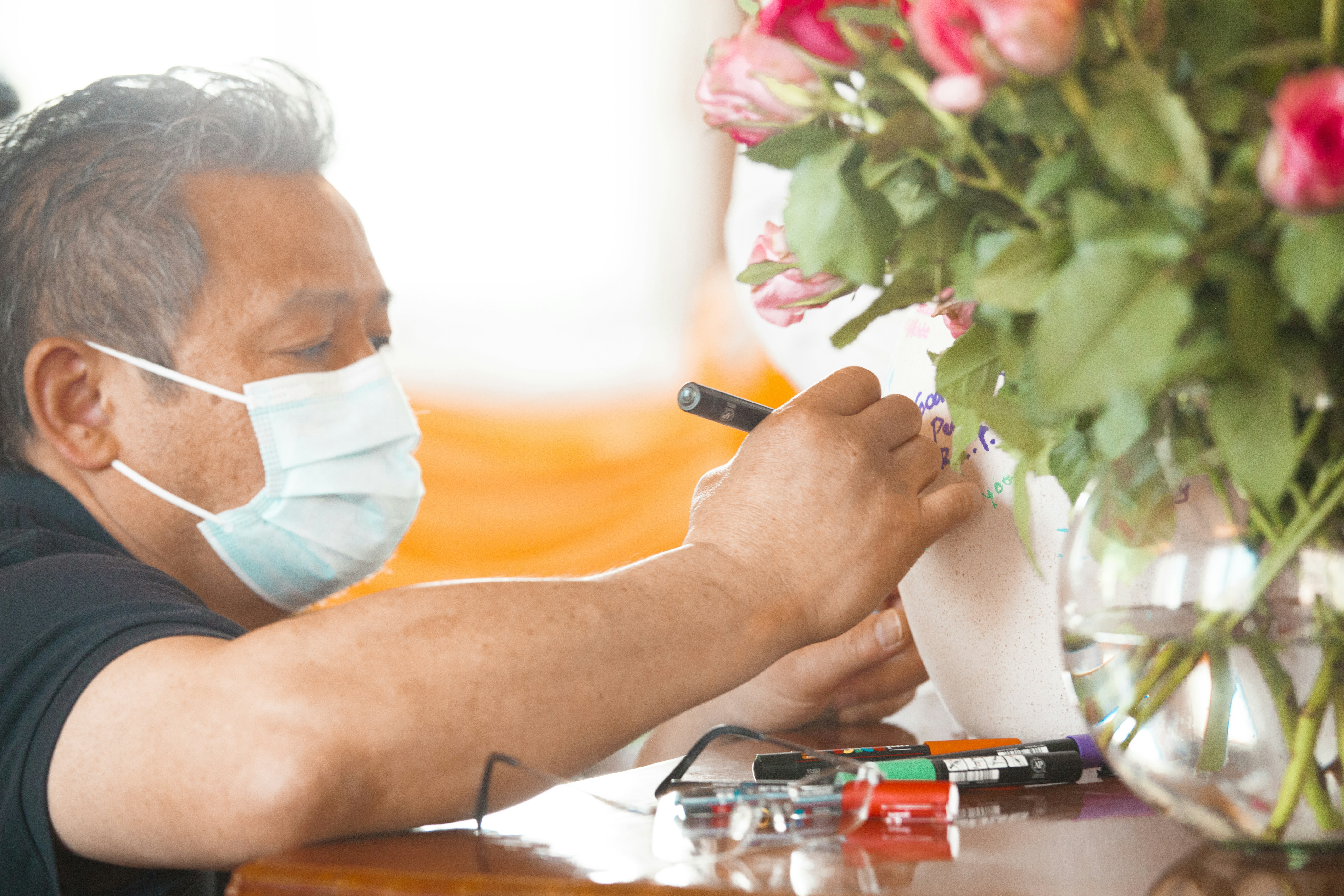 Masked Hands at the Floral Table: writing with a pencil beside pink roses
