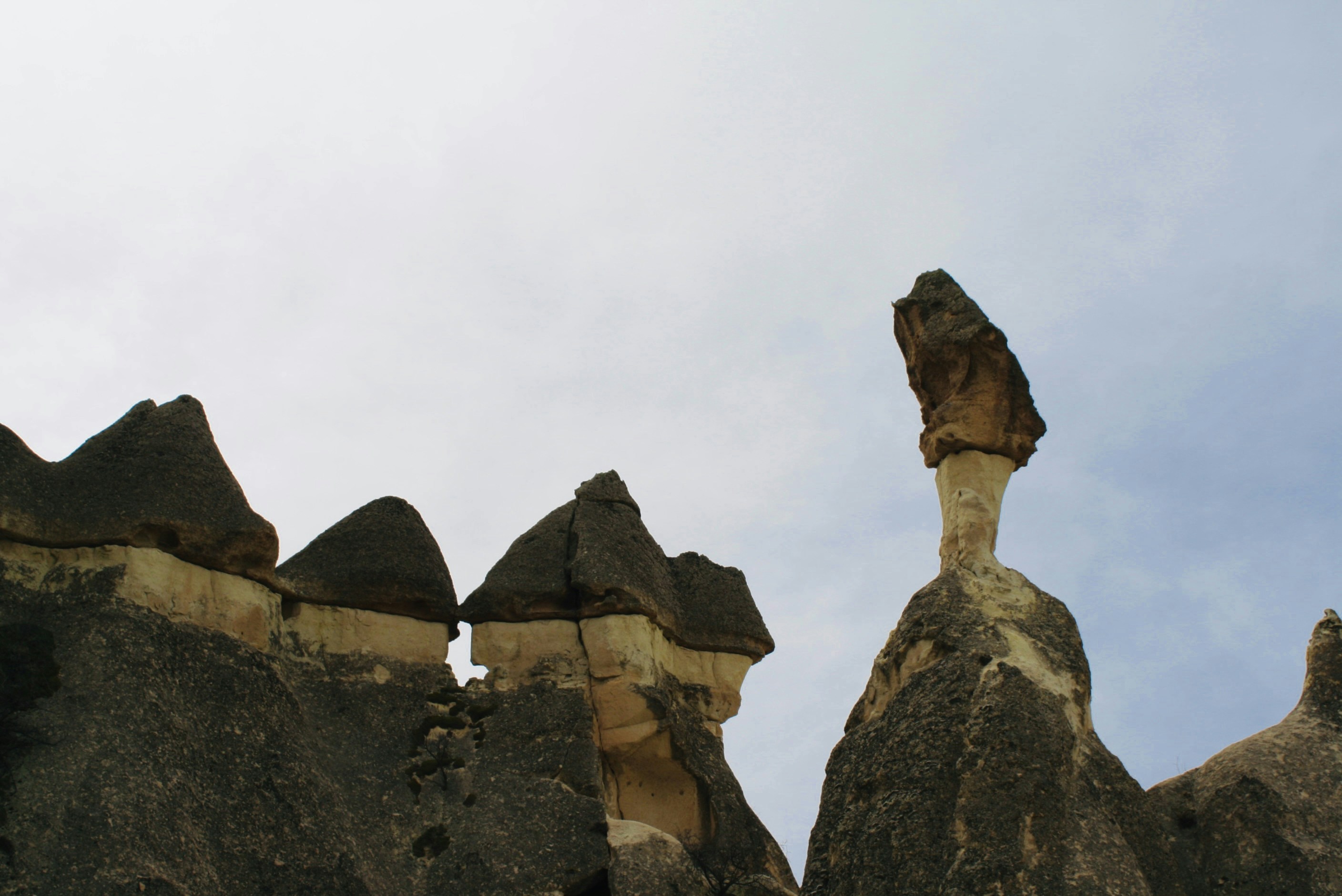 Unique rock formations rise dramatically against a cloudy sky in Cappadocia, showcasing nature's artistry in erosion. A prominent spire stands tall among the rugged landscape.