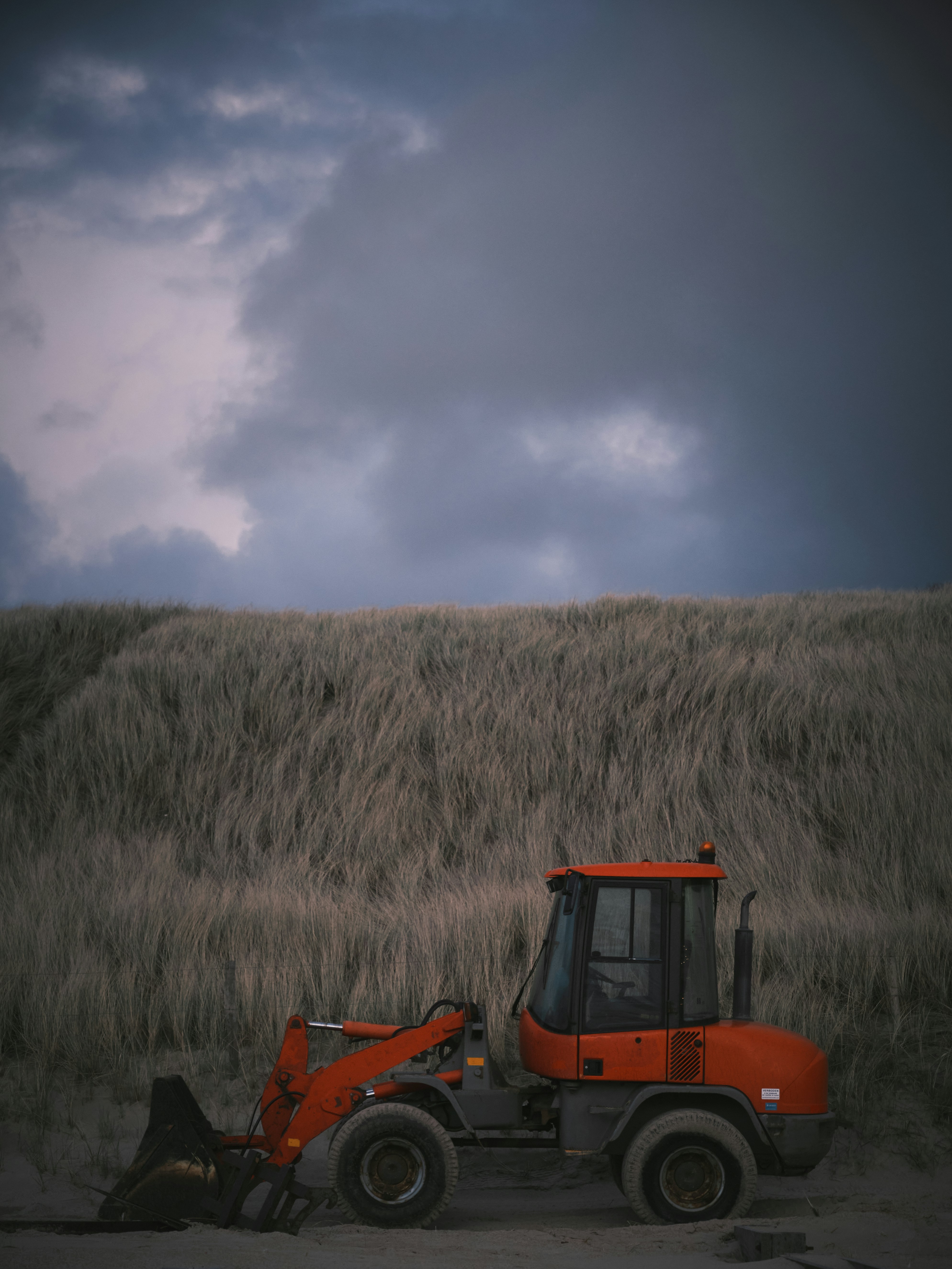 Orange construction vehicle parked against a backdrop of tall grass and moody skies.