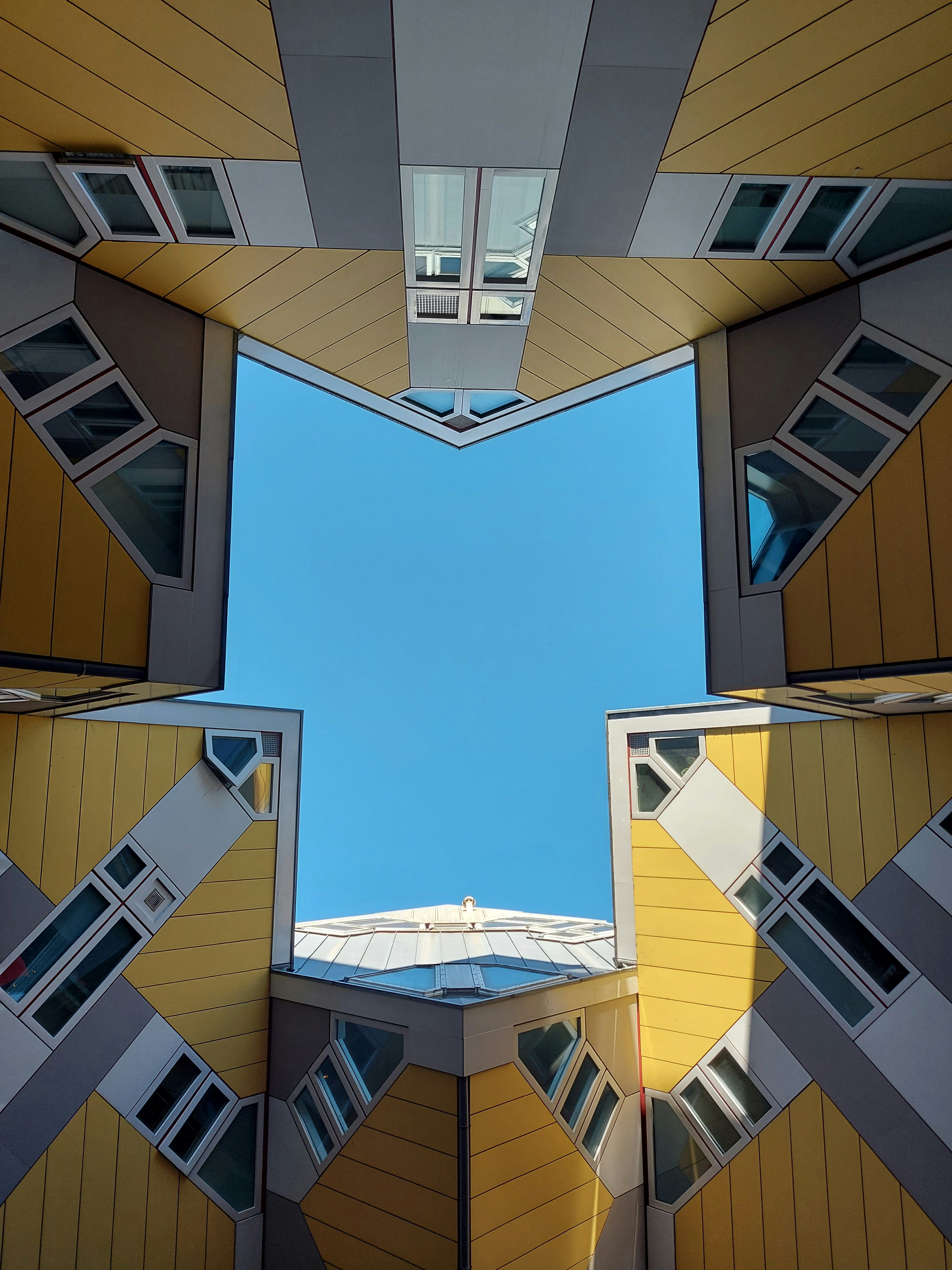 Symmetrical view from a courtyard where yellow and gray façades converge toward a central blue sky, emphasizing bold angular geometry.