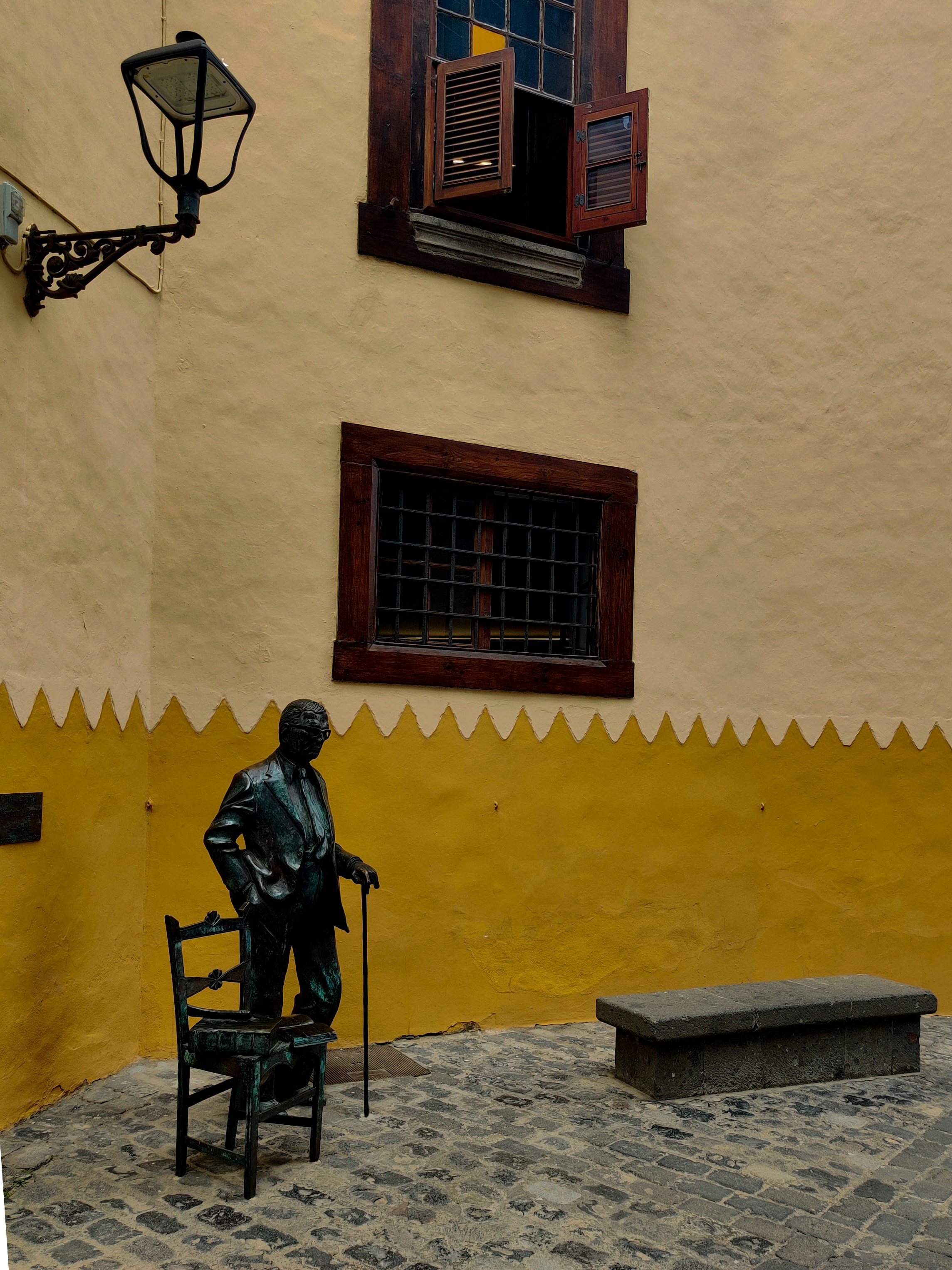Bronze statue of a distinguished figure standing beside a chair, set against a yellow wall with a window above. A vintage street lamp adds to the ambiance.