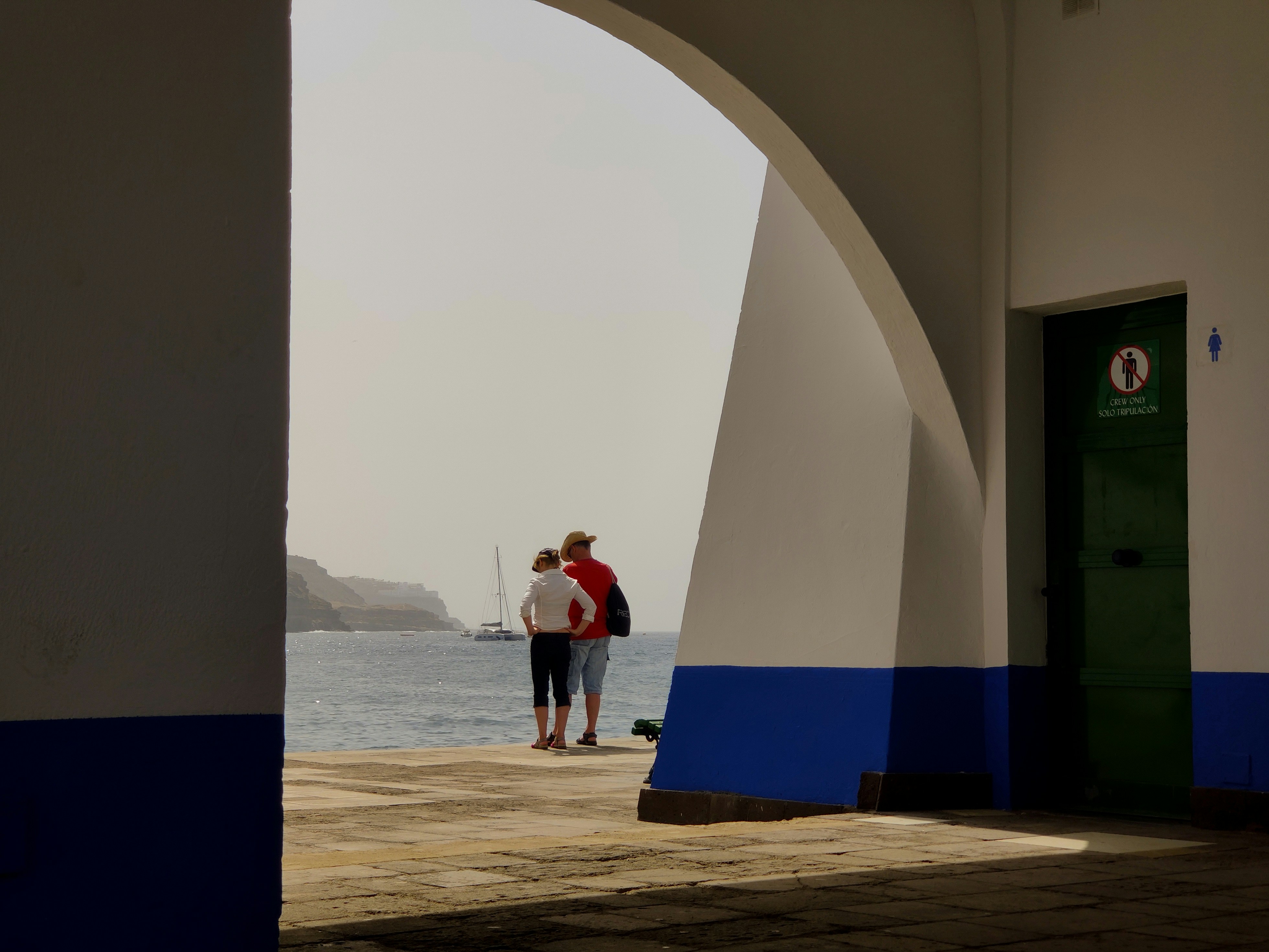 Two people stroll along a harbor beneath a white arched arcade with blue trim, sea and distant rocks in the background.