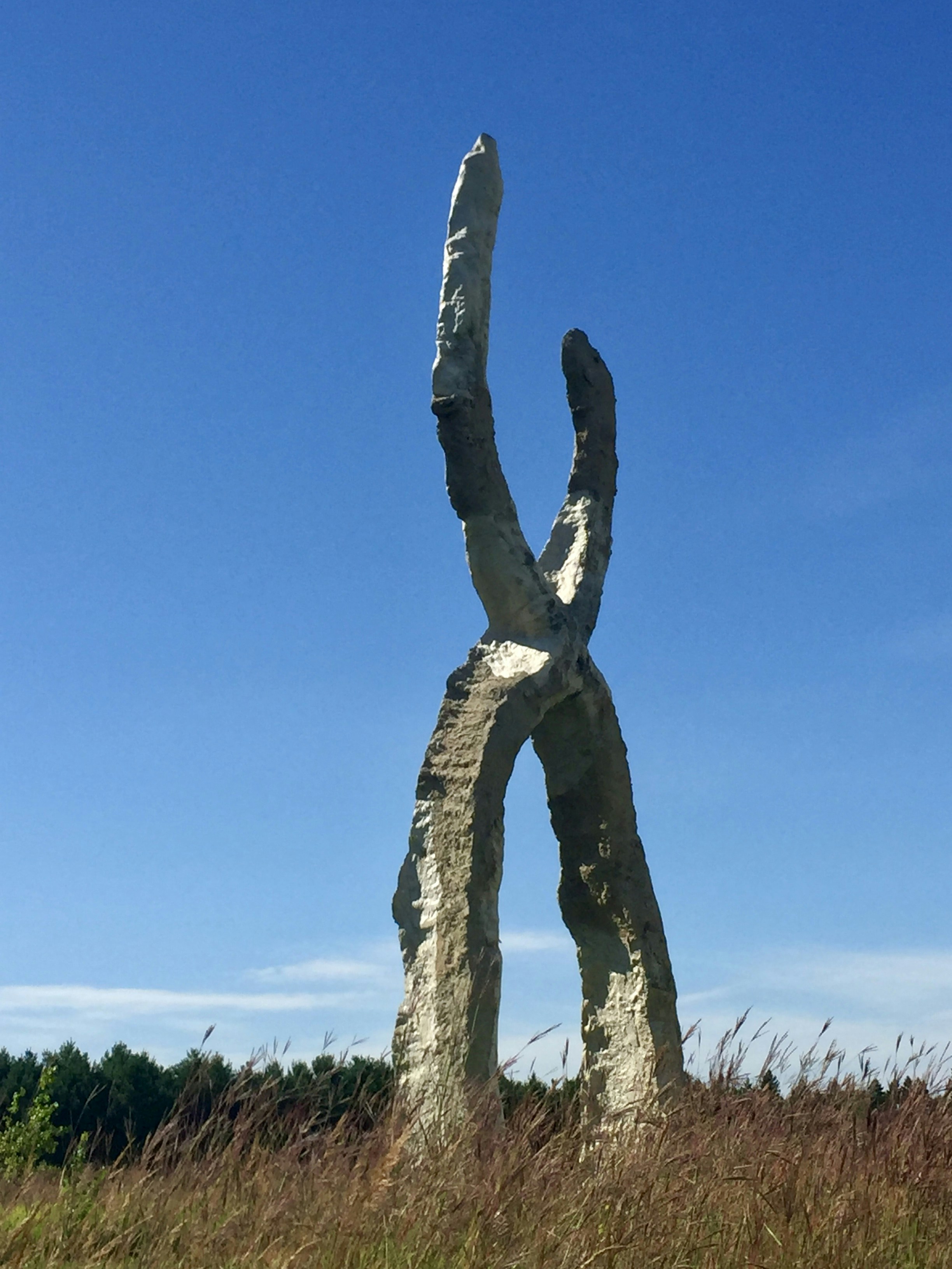 Abstract sculpture resembling open hands reaching towards the sky, set against a clear blue backdrop with tall grasses in the foreground.
