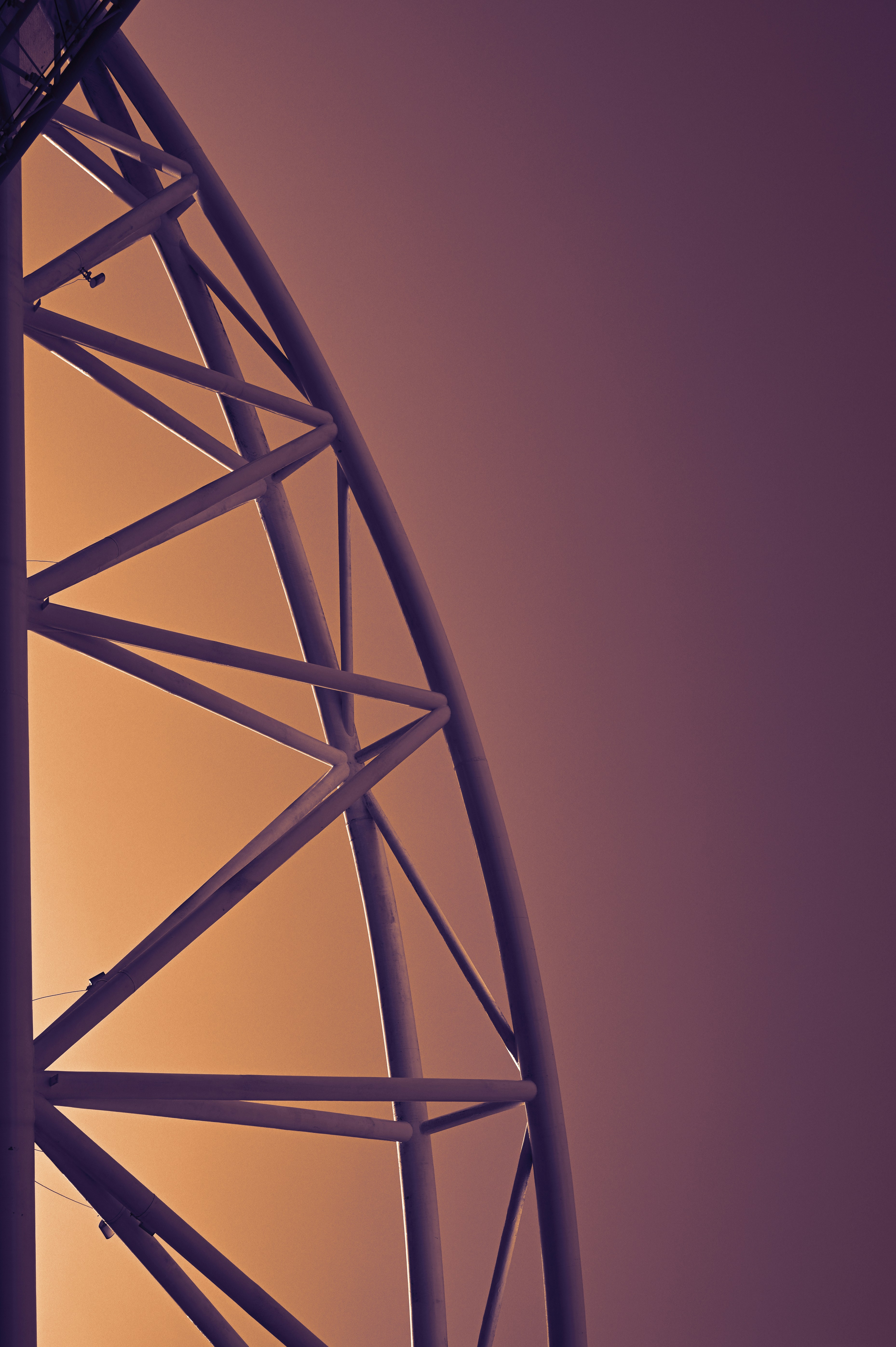 Abstract view of a Ferris wheel's structural framework against a gradient sky. The interplay of light and shadow highlights its geometric elegance.