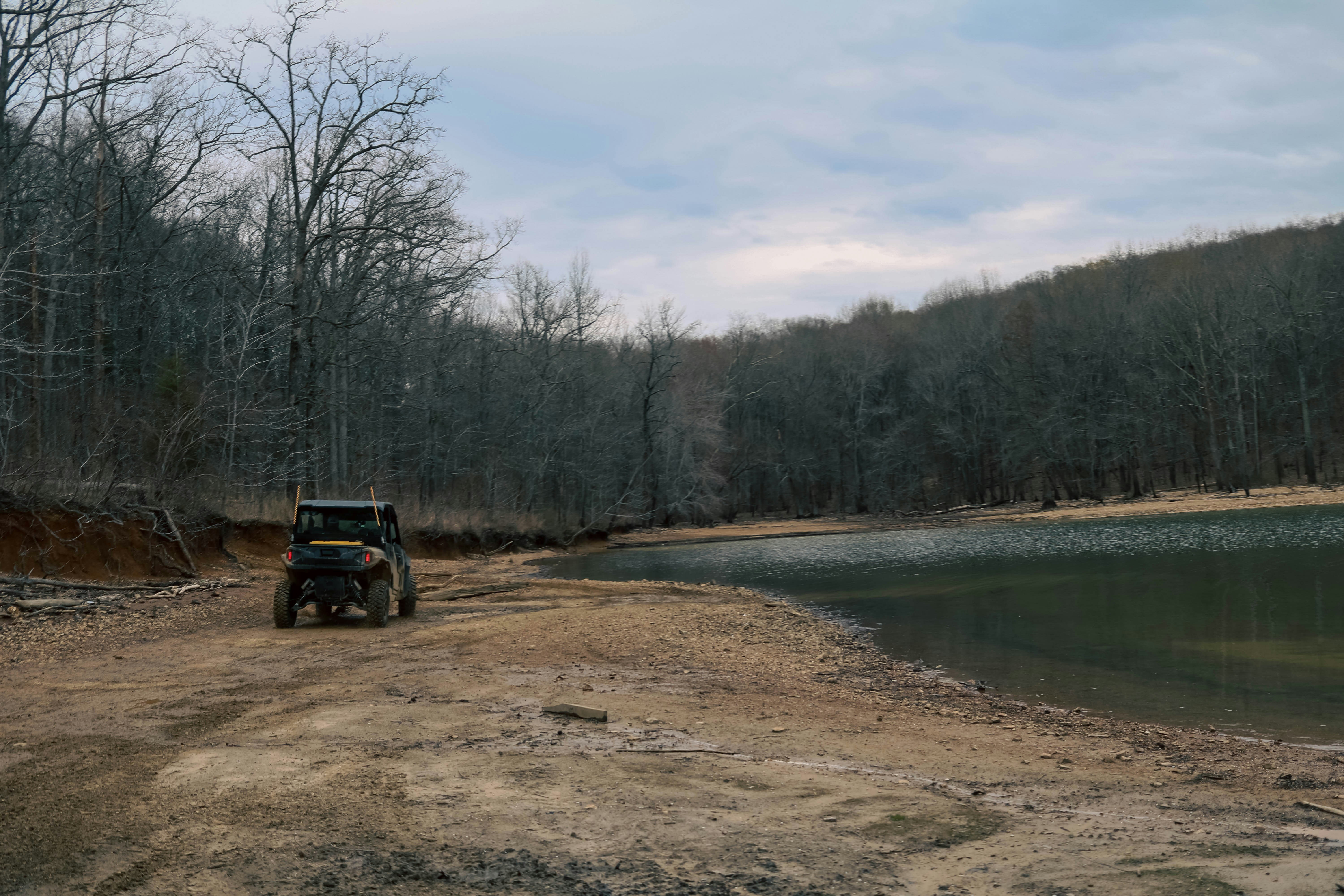 Off-road vehicle parked near a tranquil lakeside, surrounded by bare trees and a cloudy sky.