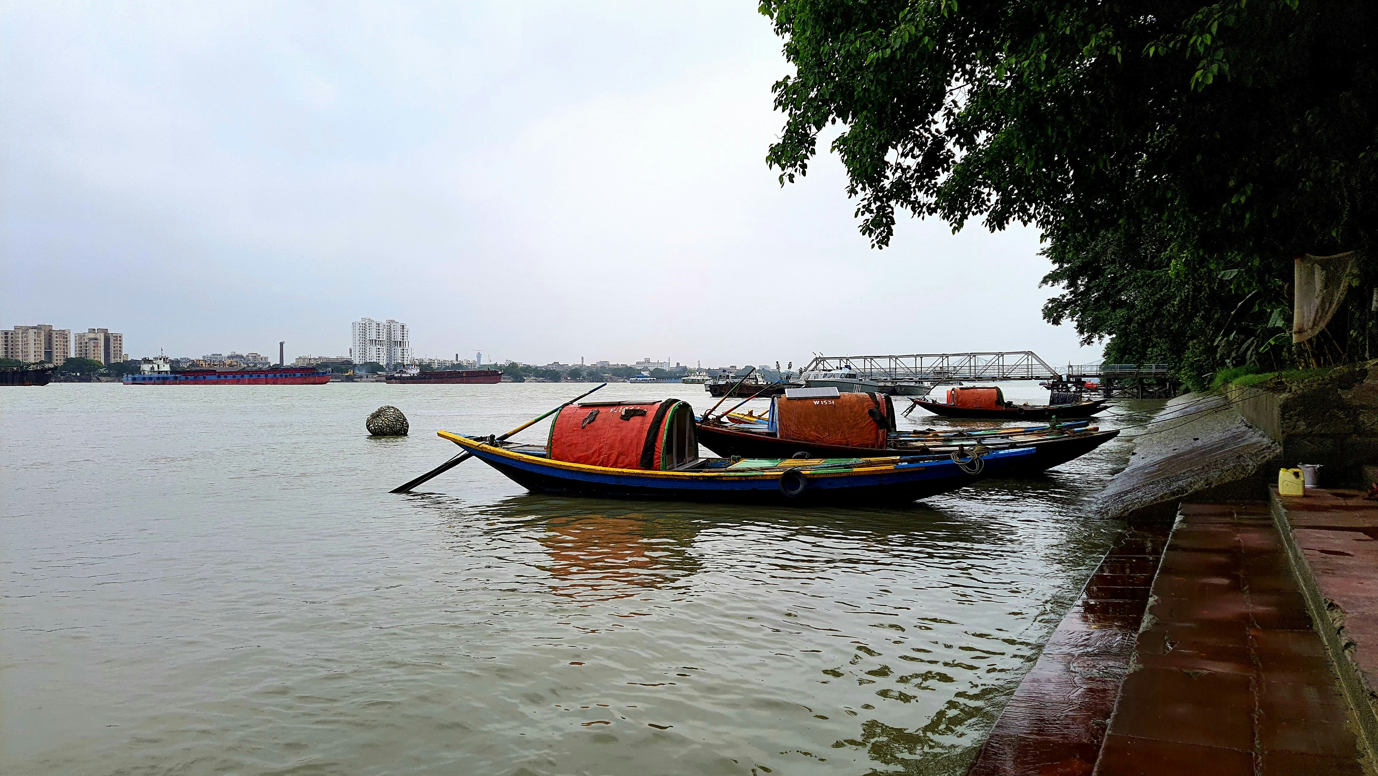 Traditional boats anchored along a riverbank, framed by a backdrop of urban buildings under a cloudy sky.