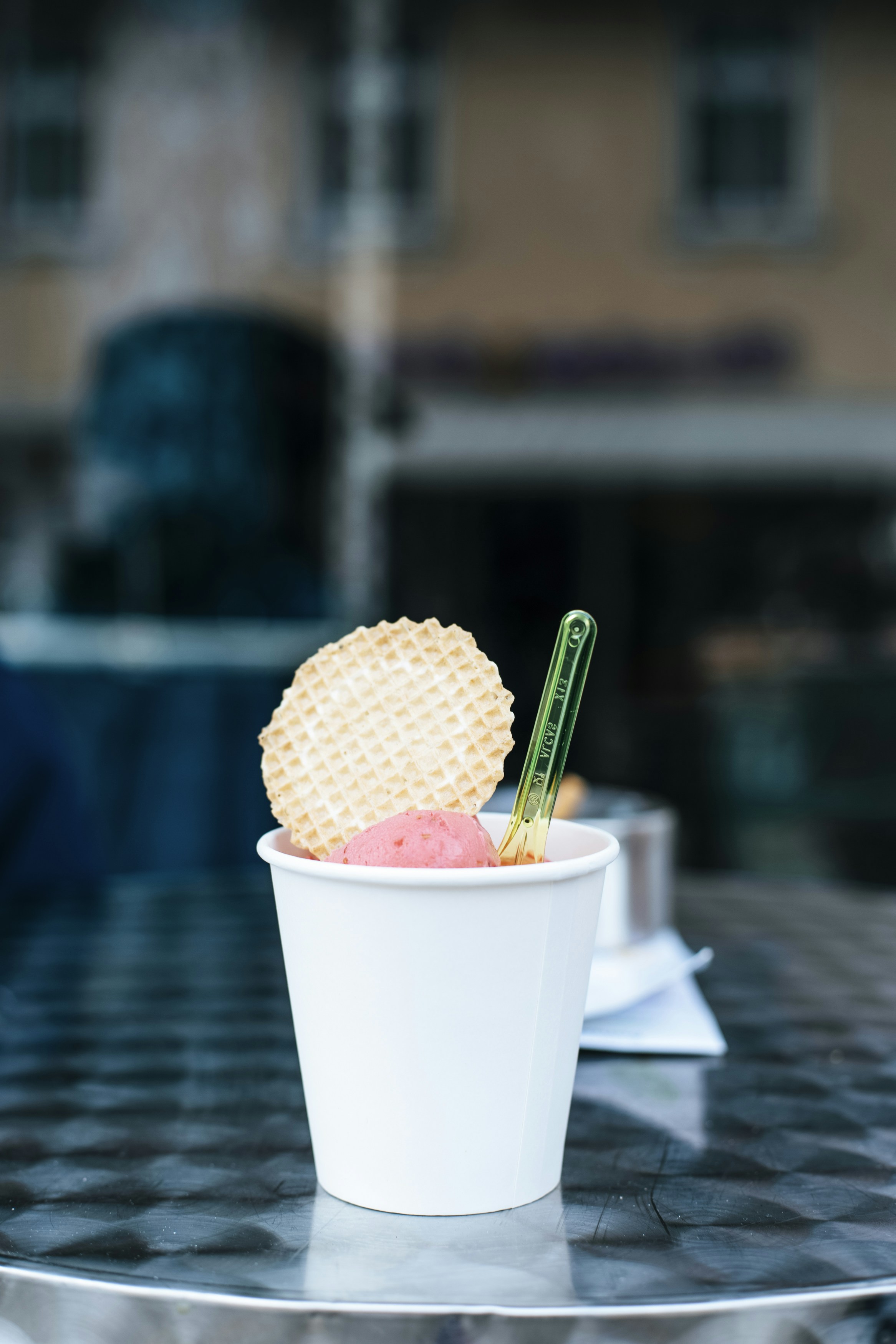 A cup of vibrant pink sorbet topped with a waffle cone and a green spoon, set against a blurred urban backdrop.