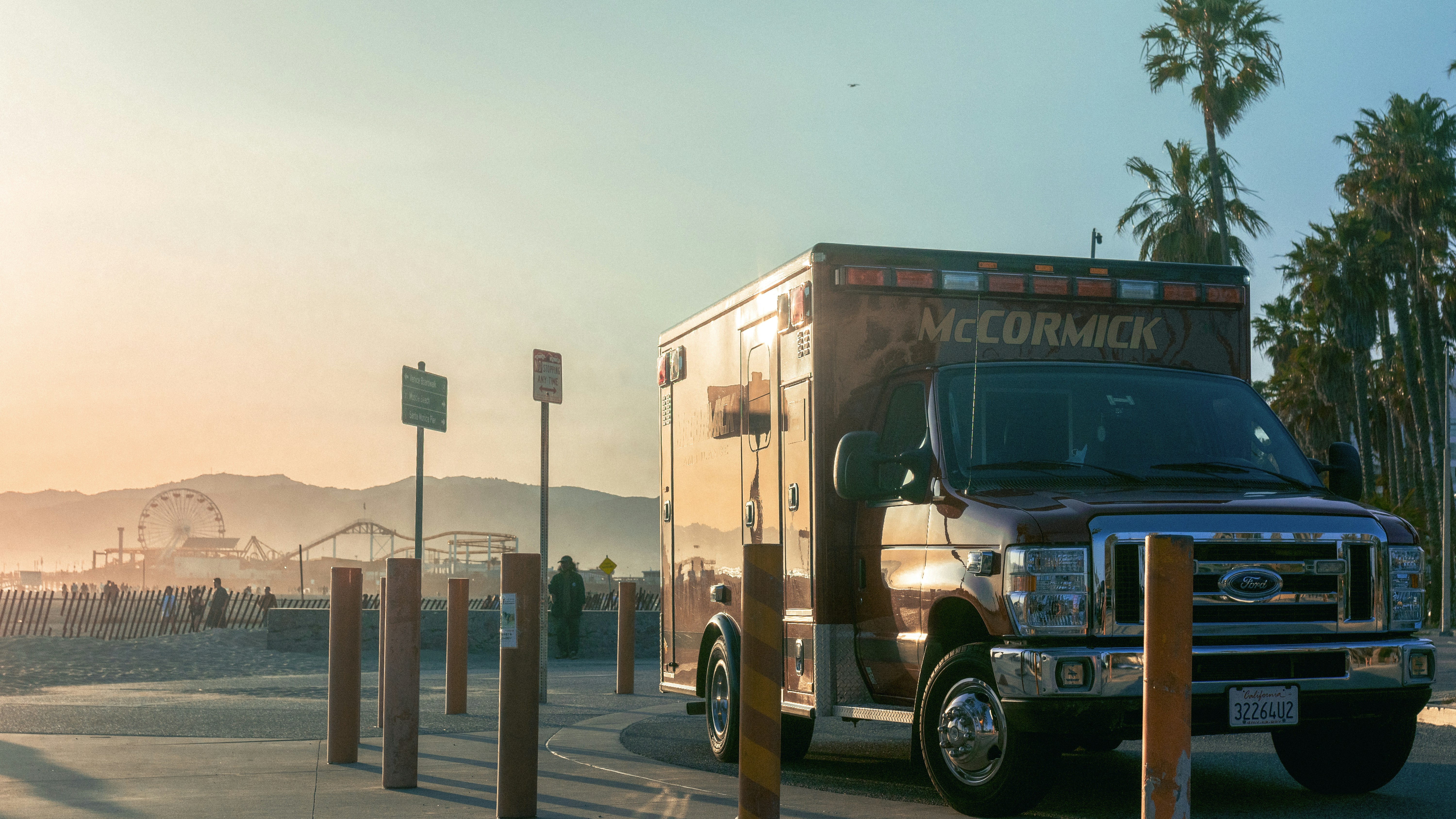 Ambulance parked along the beach with palm trees and a distant amusement park in the background, capturing a moment of calm amidst the hustle of the coast.