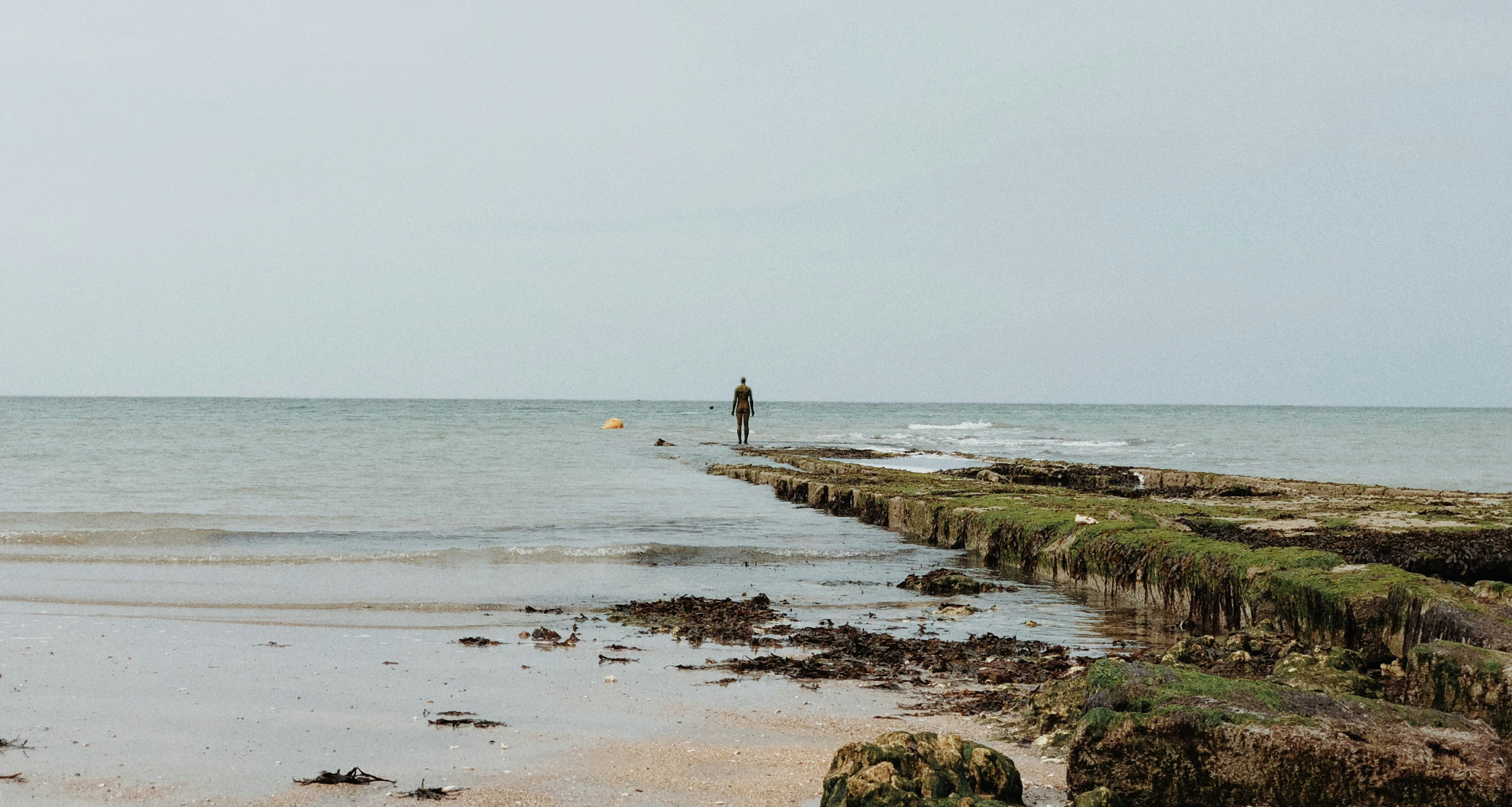Person standing on a rocky pier extending into the calm sea under an overcast sky.