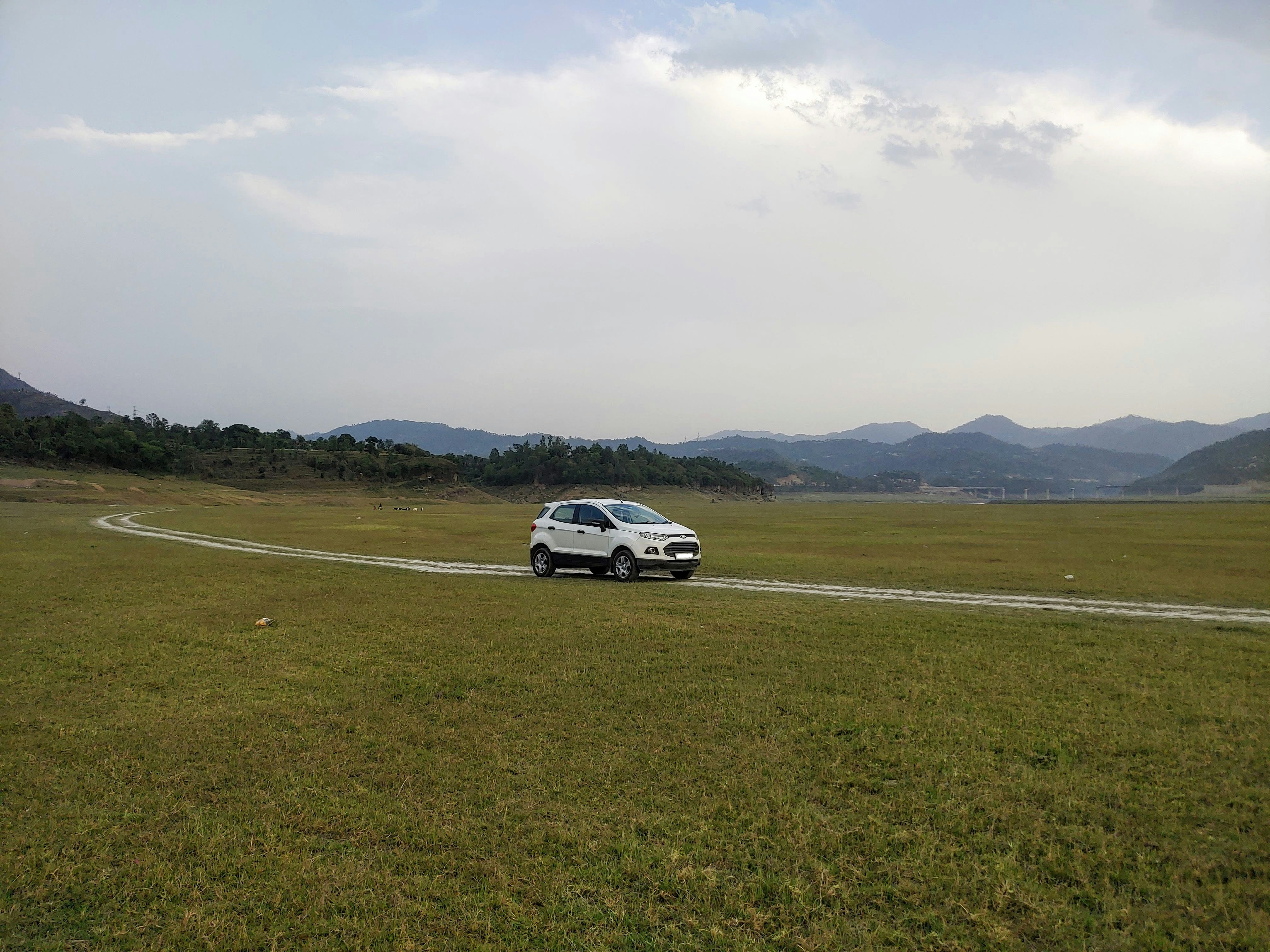 White car traveling on a winding road through expansive green fields with distant hills under a cloudy sky.