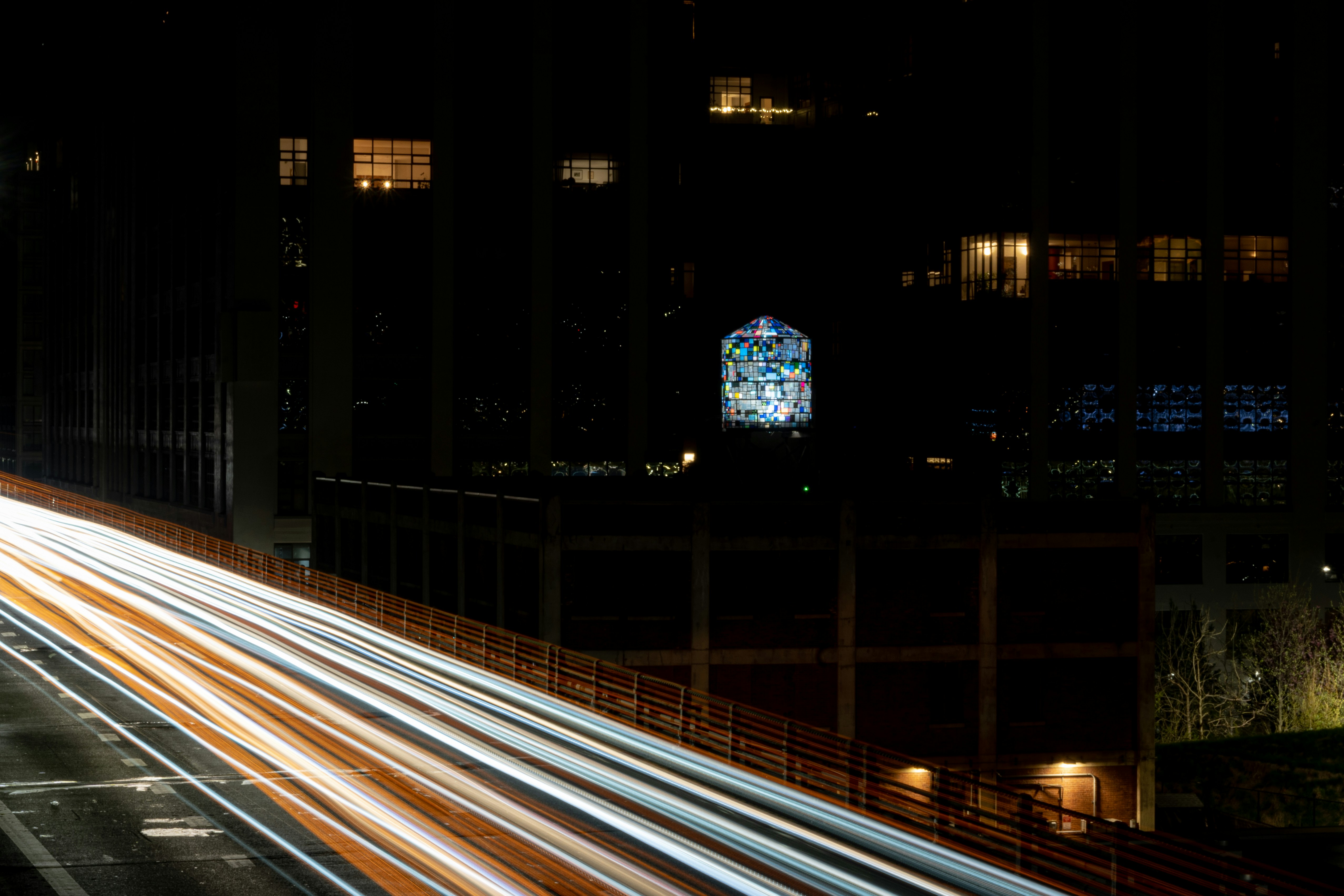 A vibrant display of light trails from passing vehicles contrasts against a distant illuminated window, creating a dynamic urban scene at night.