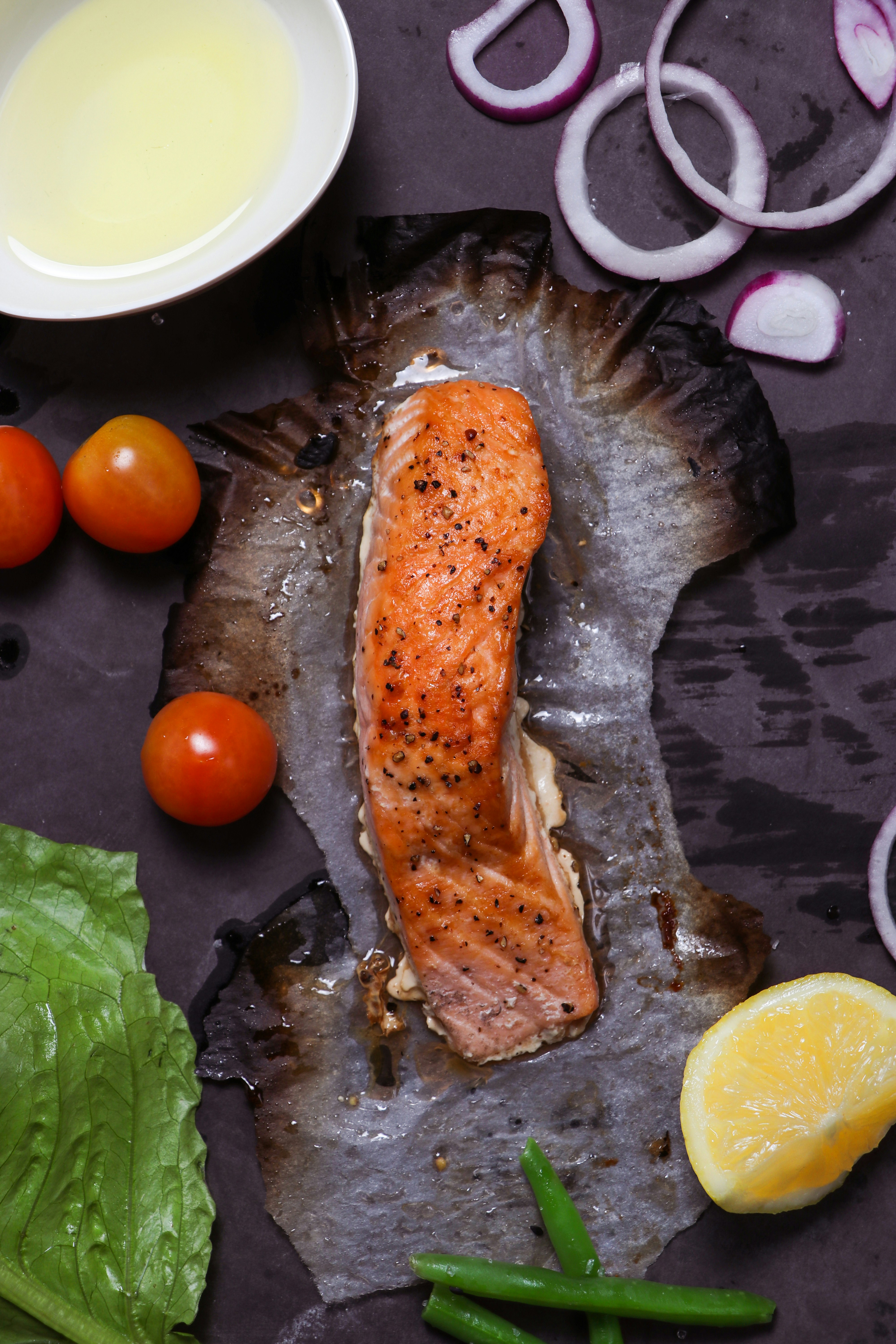 Grilled salmon fillet resting on parchment paper, surrounded by cherry tomatoes, lettuce, green beans, and lemon slices.