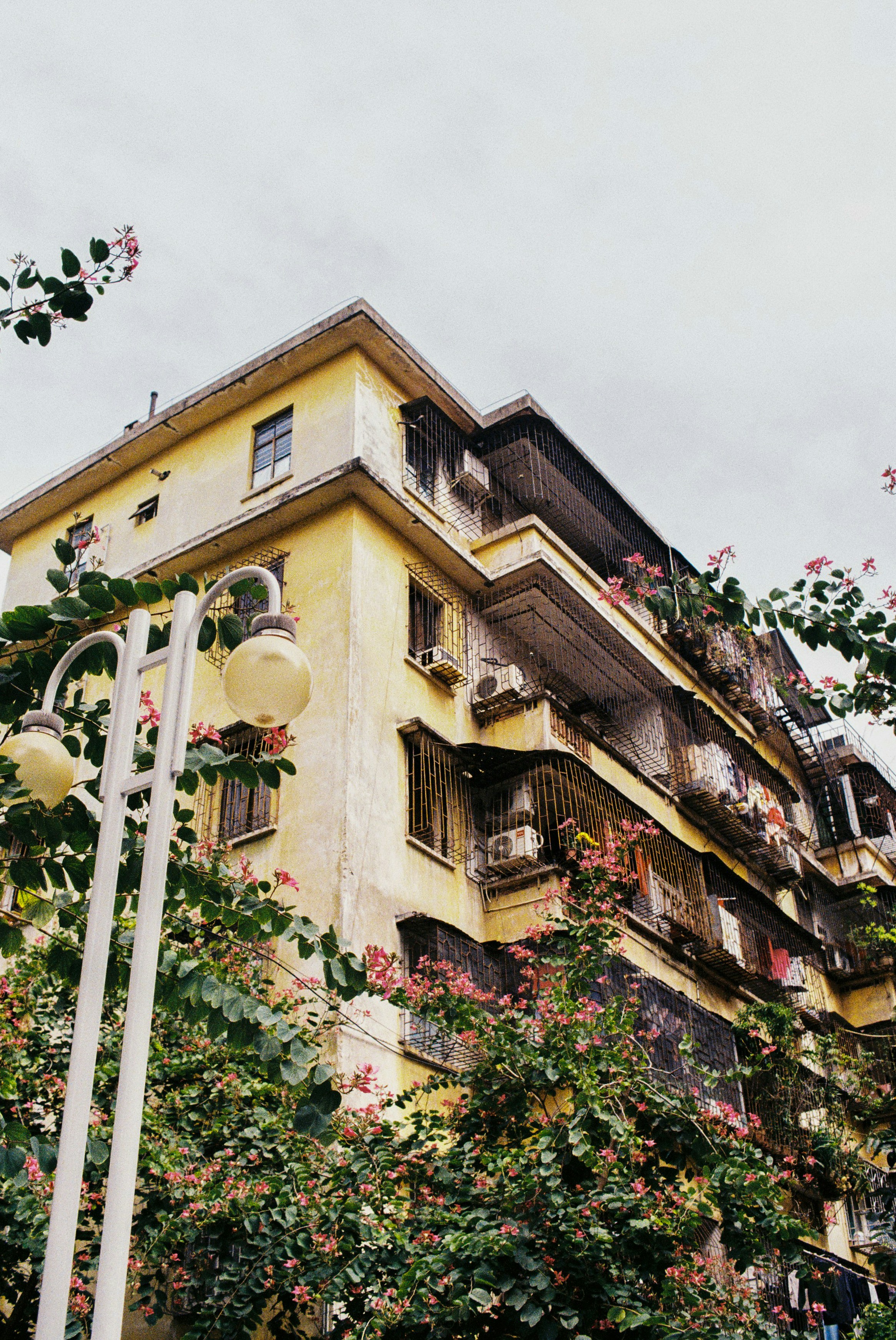 Yellow building with balconies surrounded by flowering plants and vintage streetlights under a cloudy sky.