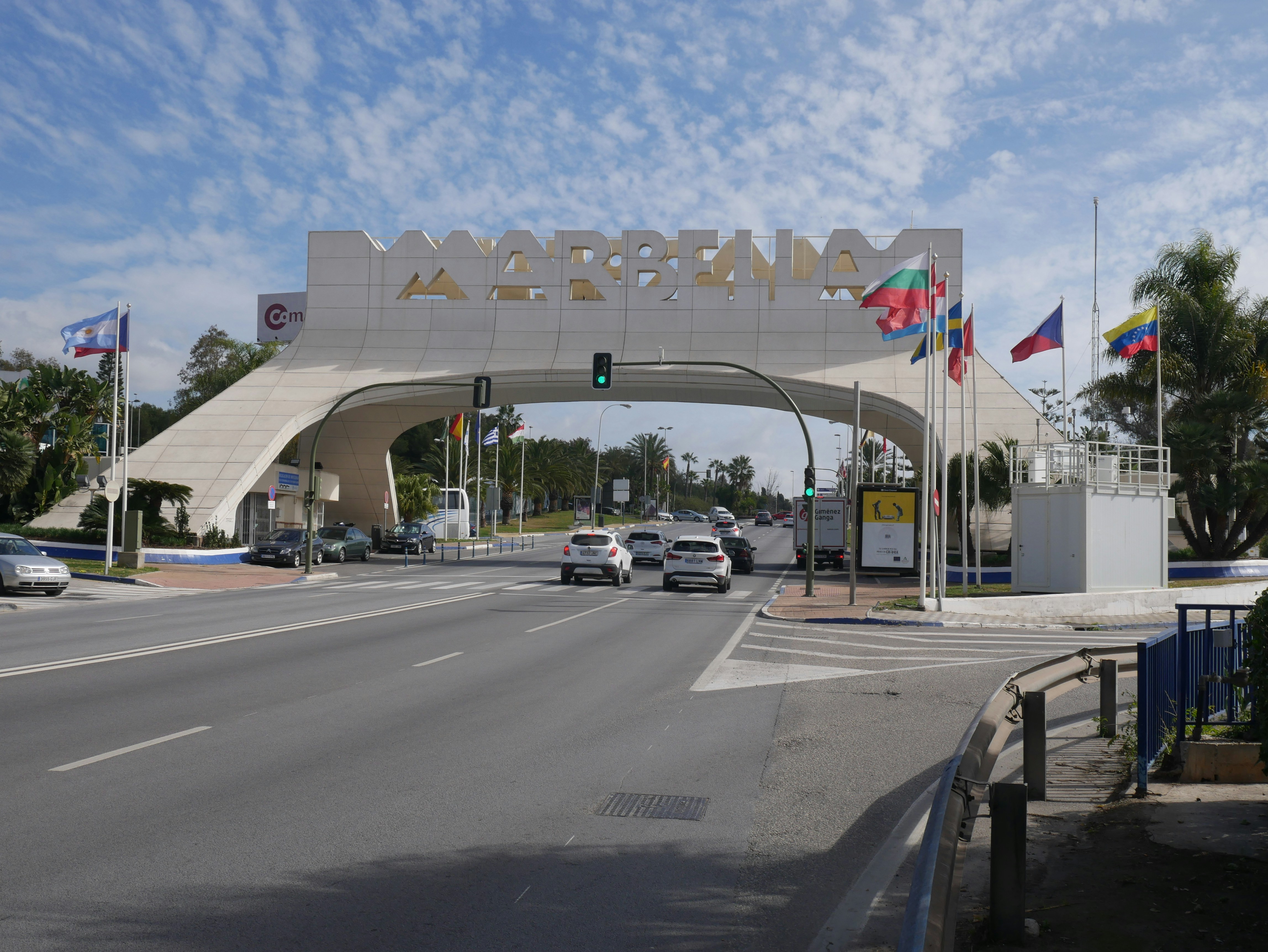 Cars approach an archway adorned with international flags under a blue sky.