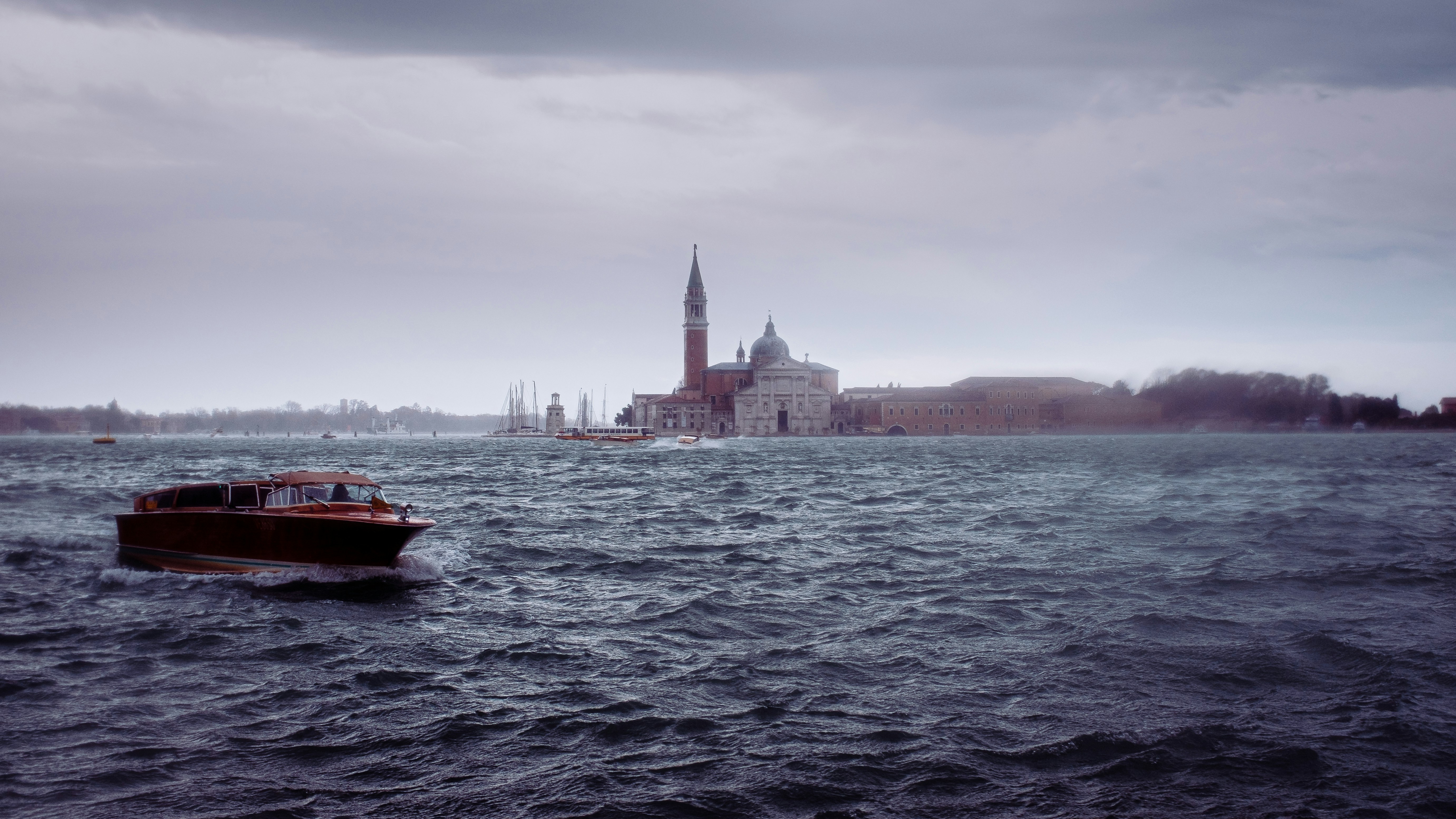 A wooden boat navigates choppy waters near a distant Venetian skyline under an overcast sky.