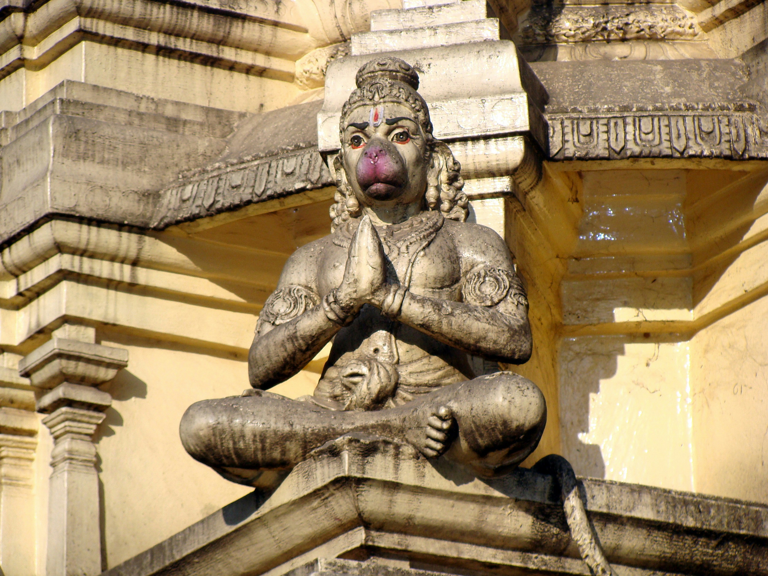 Stone Hanuman statue seated in lotus position with hands in prayer inside a sunlit temple alcove.