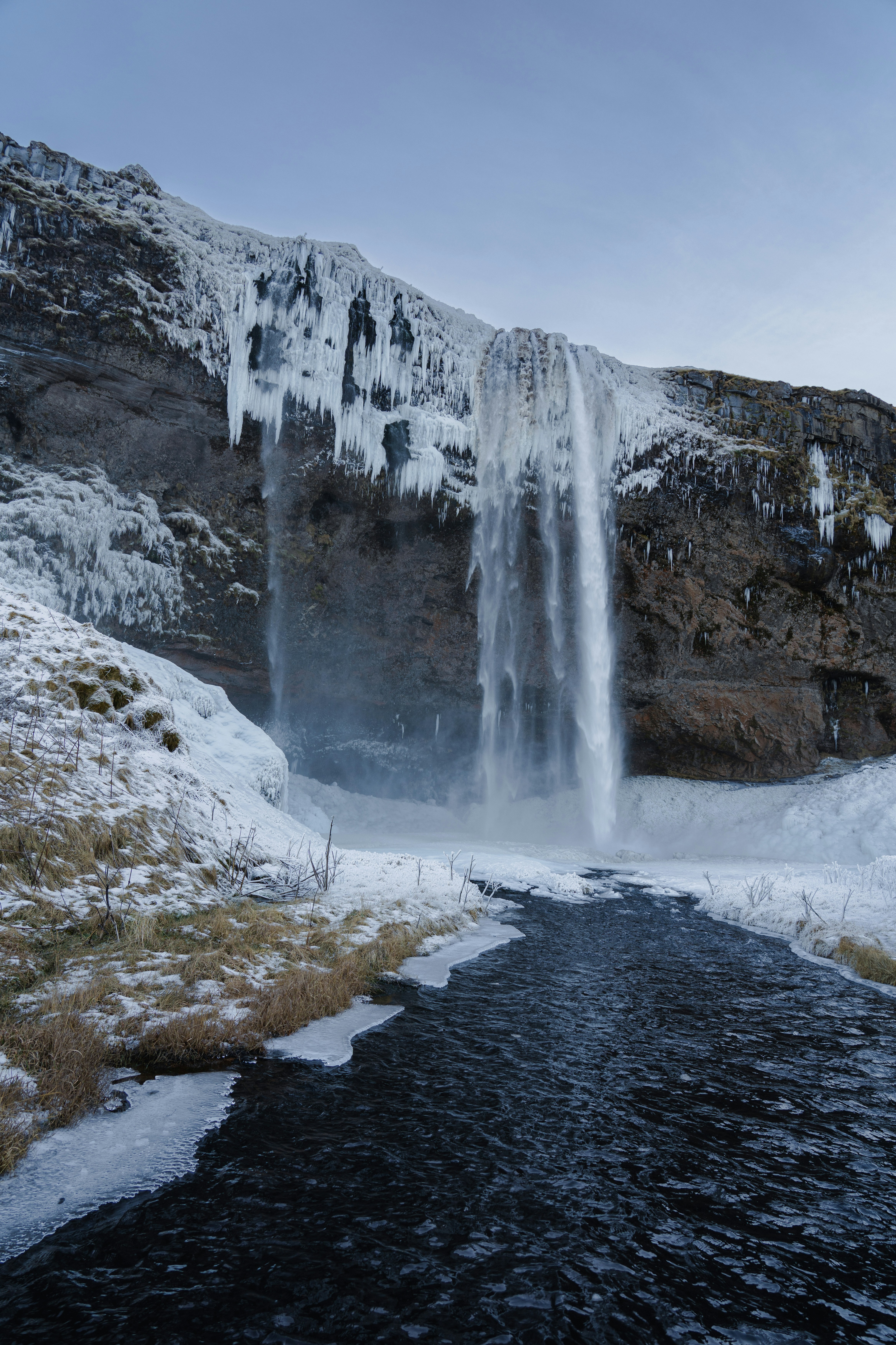 Majestic waterfall cascading down a cliff, surrounded by icy formations and a serene river. The scene captures the raw beauty of winter's embrace.