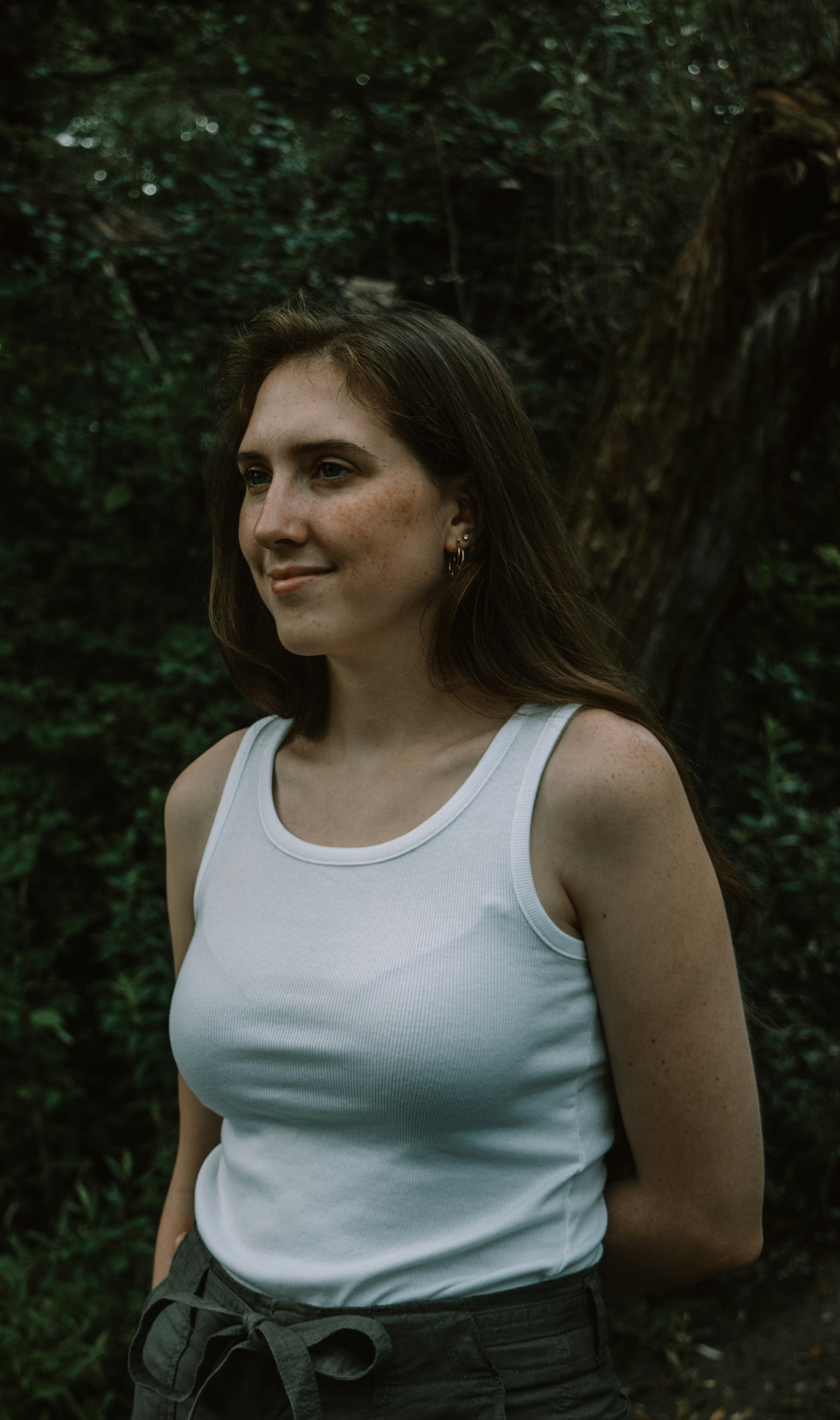 Young woman in a white tank top stands against a lush green backdrop, exuding a serene presence. The natural setting enhances her relaxed demeanor.
