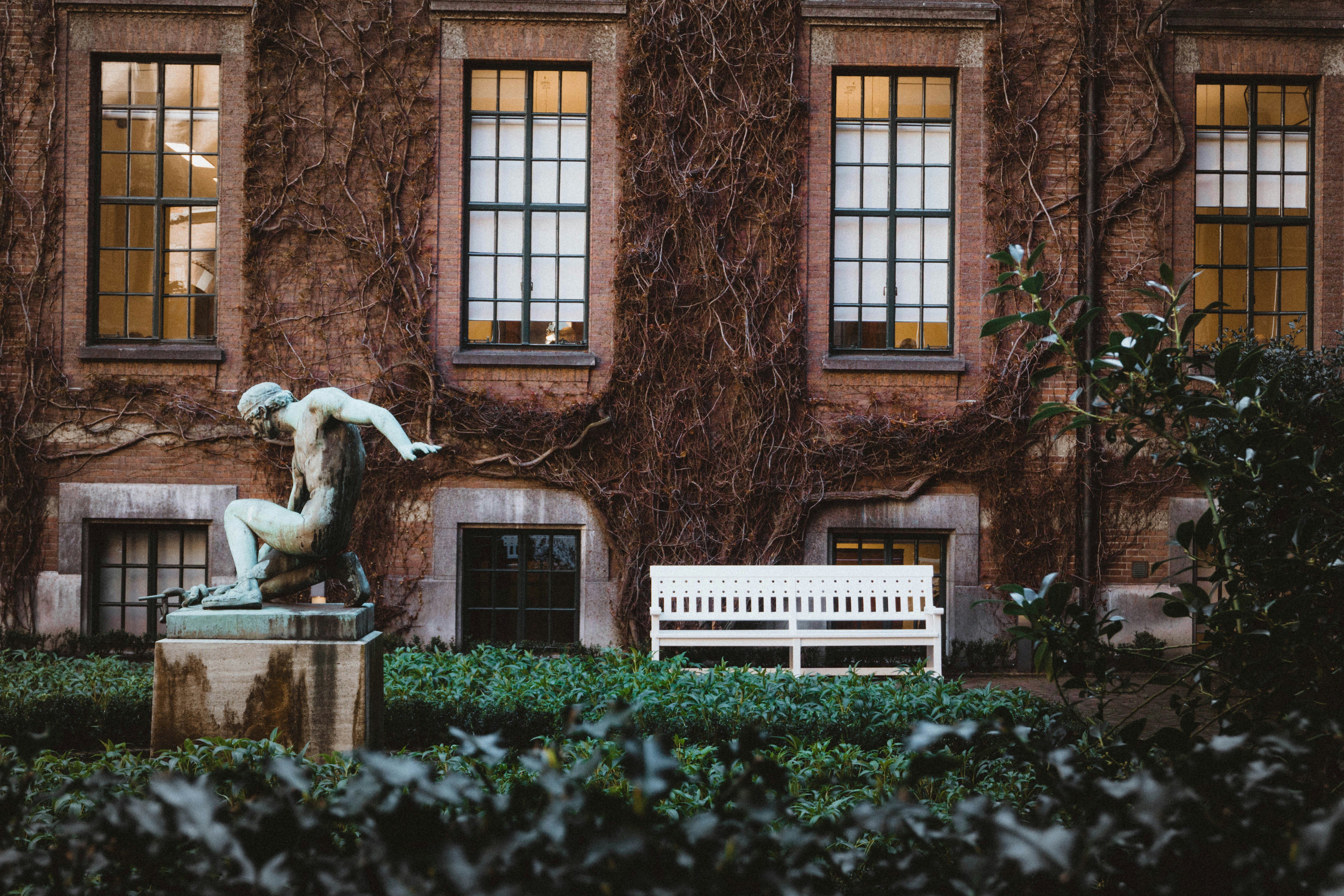 Bronze sculpture of a figure in a contemplative pose, surrounded by lush greenery and an ivy-covered brick wall. A white bench adds to the tranquil atmosphere.