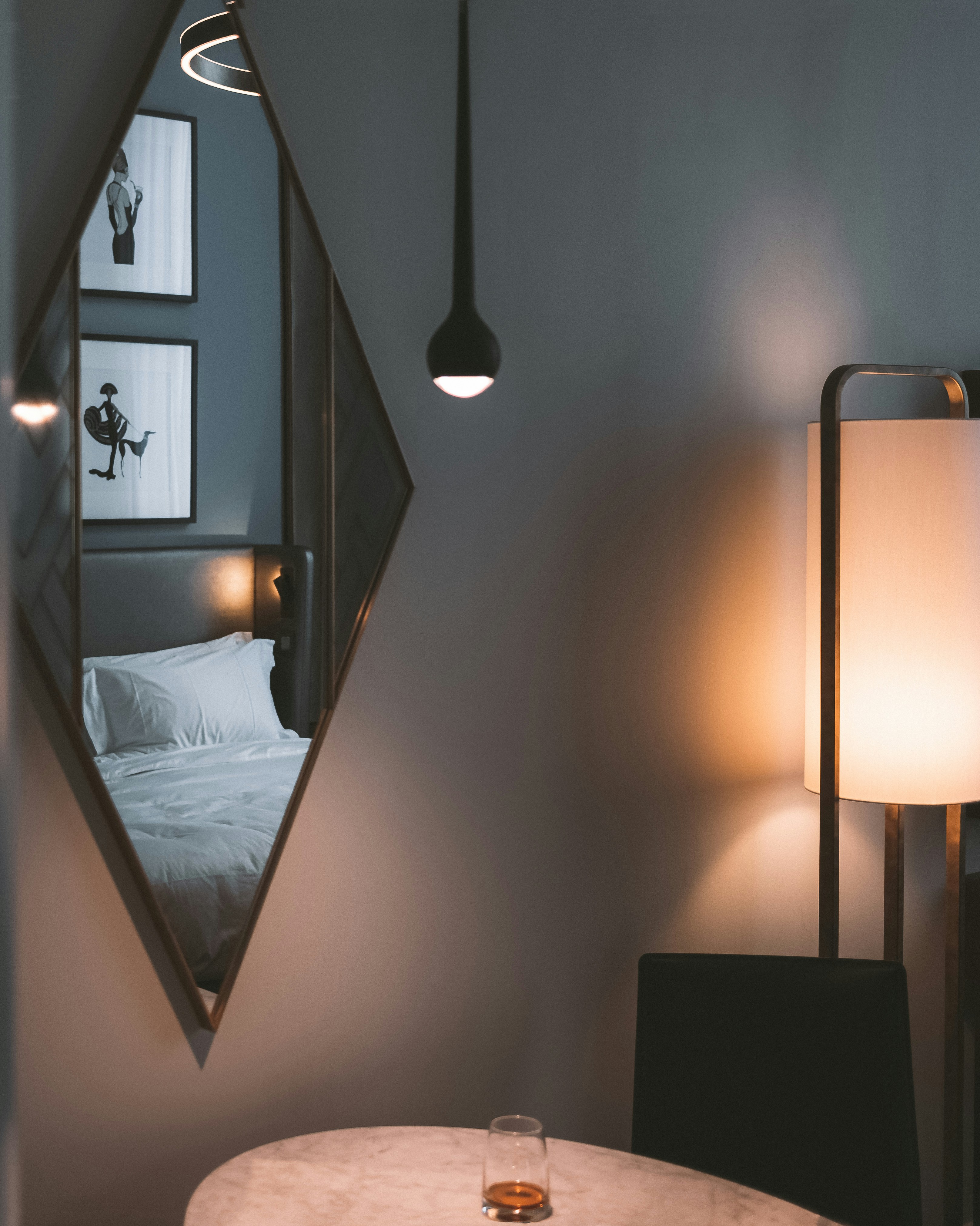A stylish interior featuring a geometric mirror reflecting a cozy bed and framed artwork, complemented by warm lighting from a modern lamp.