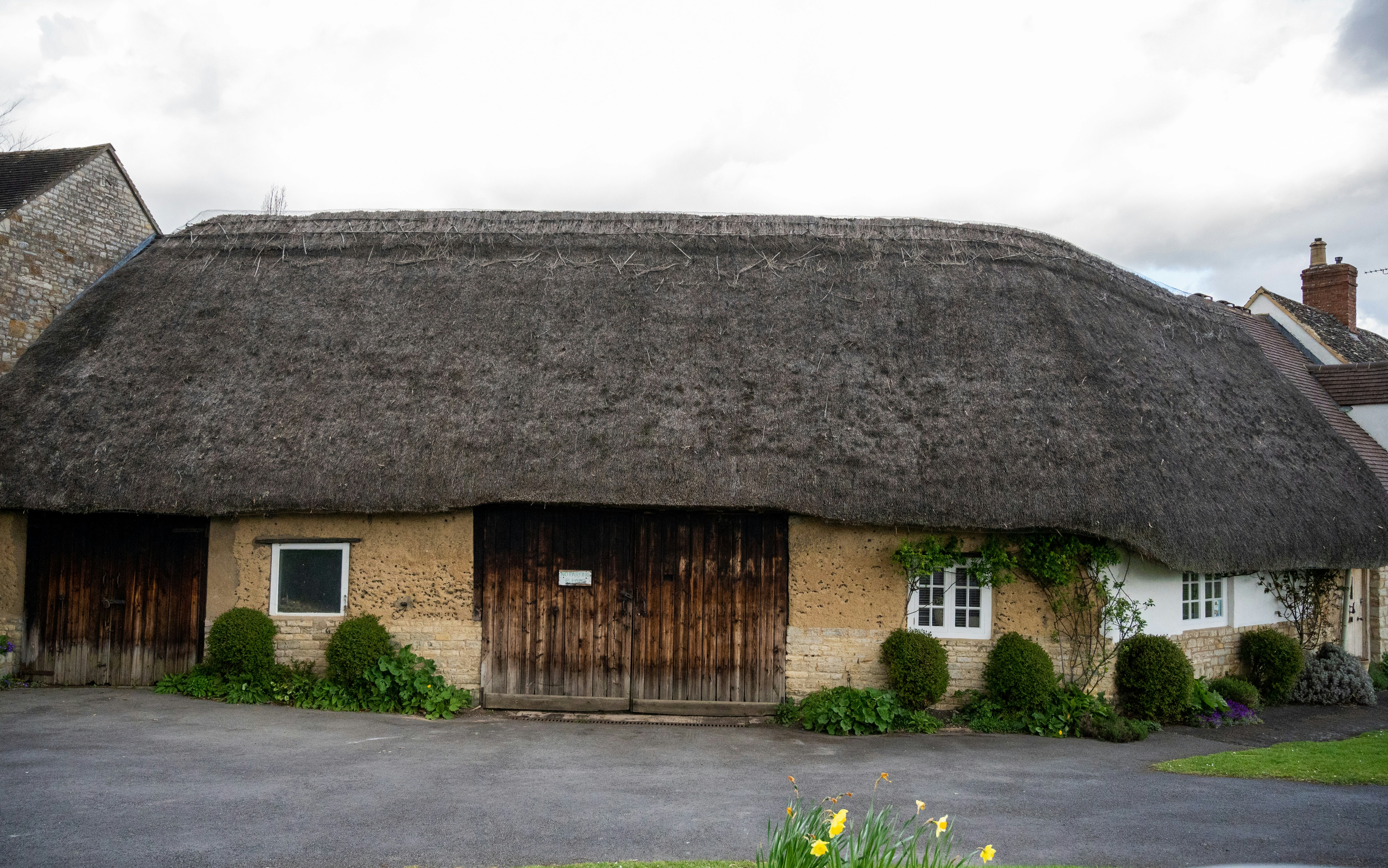 Traditional thatched-roof cottage with wooden doors and lush greenery surrounding the entrance. The structure reflects classic architectural style.