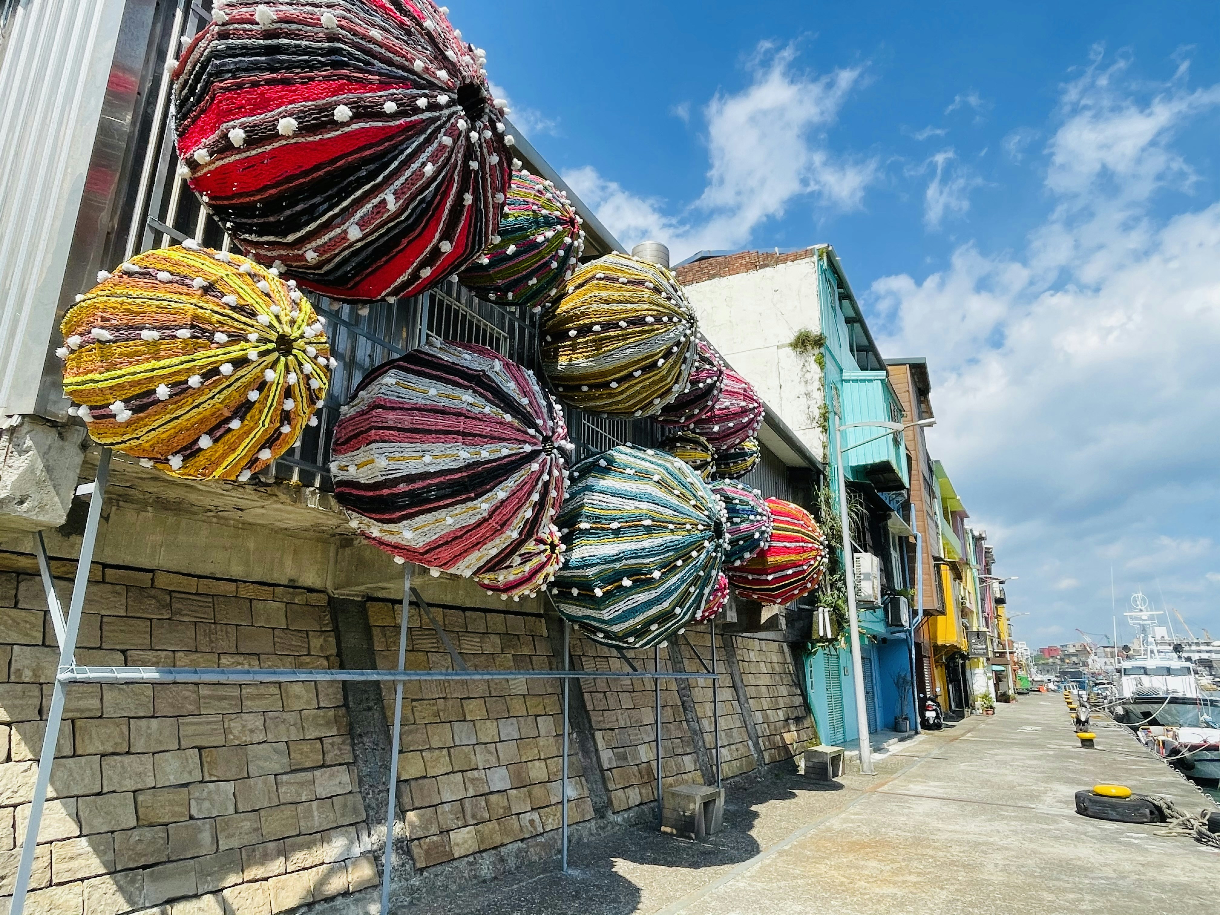 Colorful, oversized decorative canopies hang from a waterfront building, creating a vibrant visual contrast against the blue sky and stone wall.
