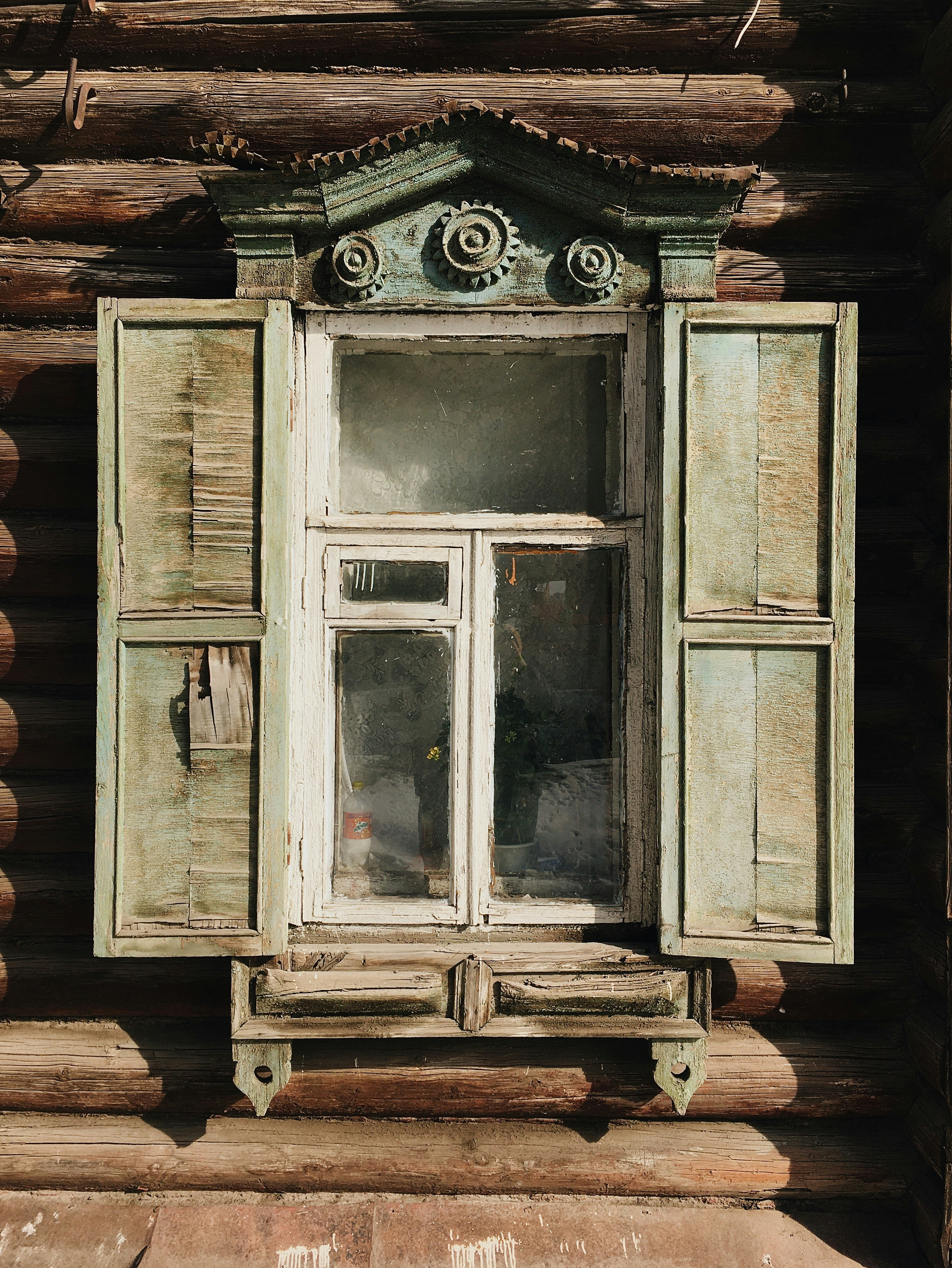 A vintage wooden window with ornate shutters, showcasing intricate carvings and a faded green hue against a rustic log wall.