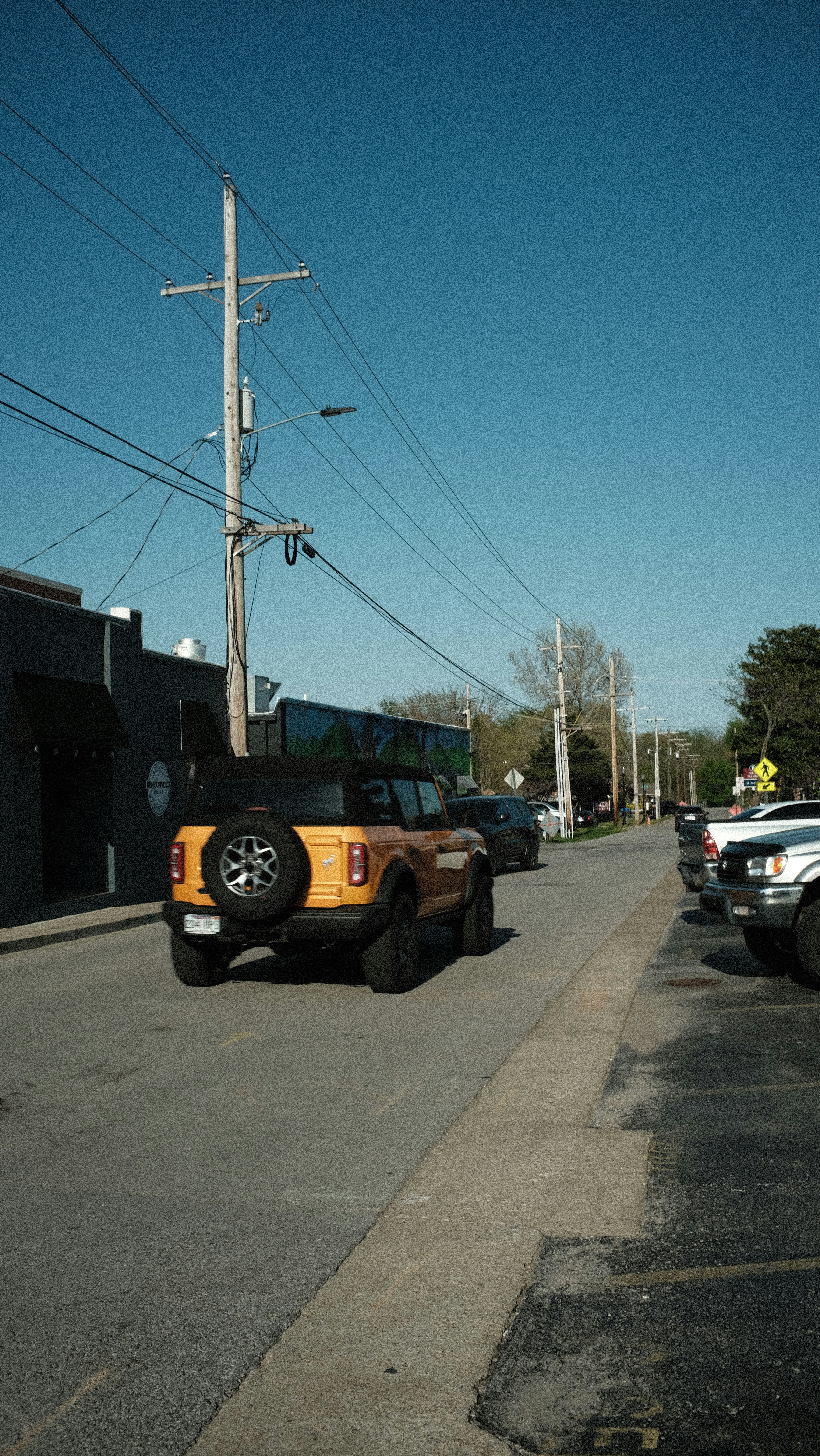 A yellow car driving down the street past power lines.