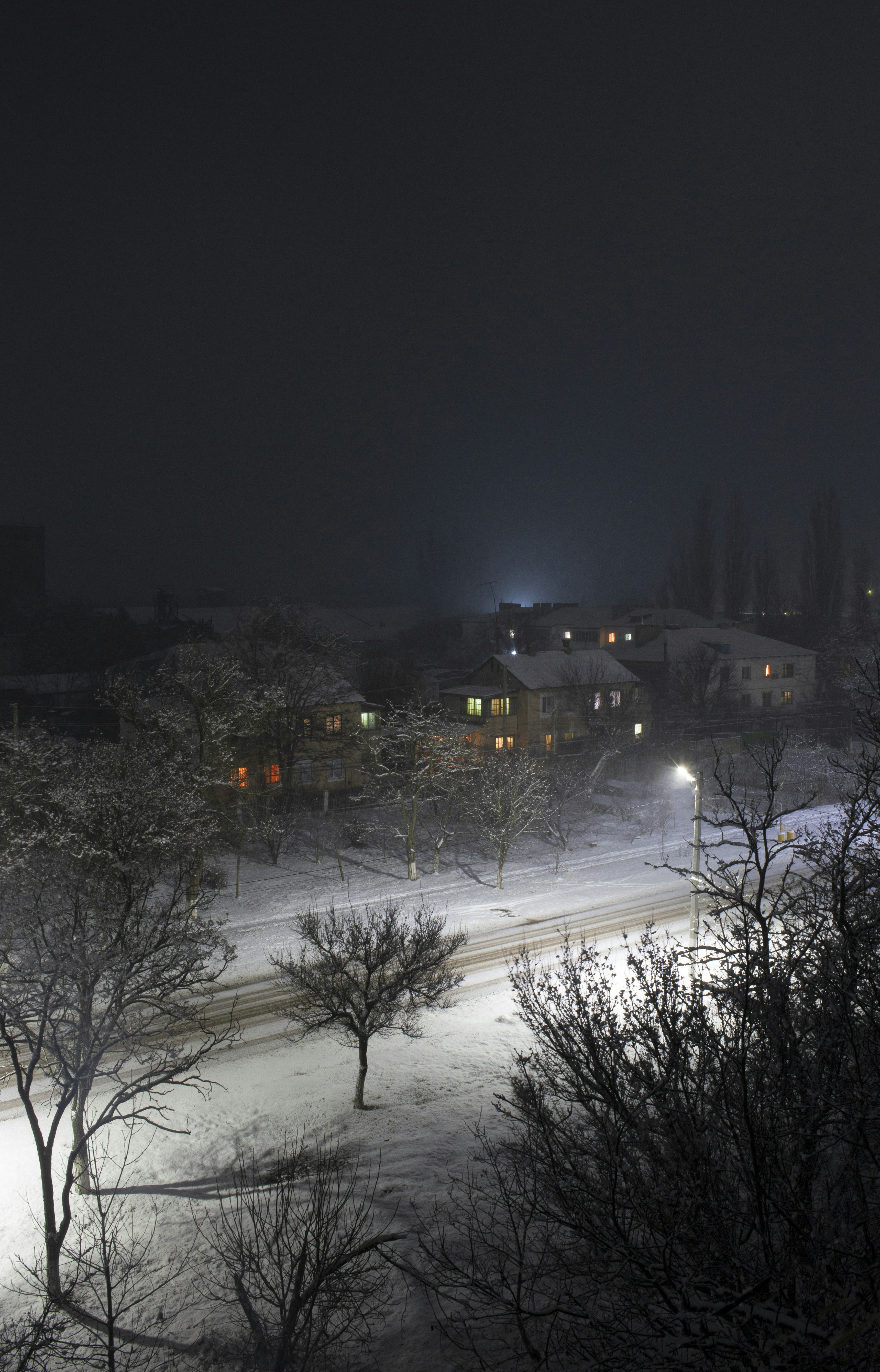 Snow-covered street illuminated by streetlights, with warm windows glowing in nearby houses during a winter night.