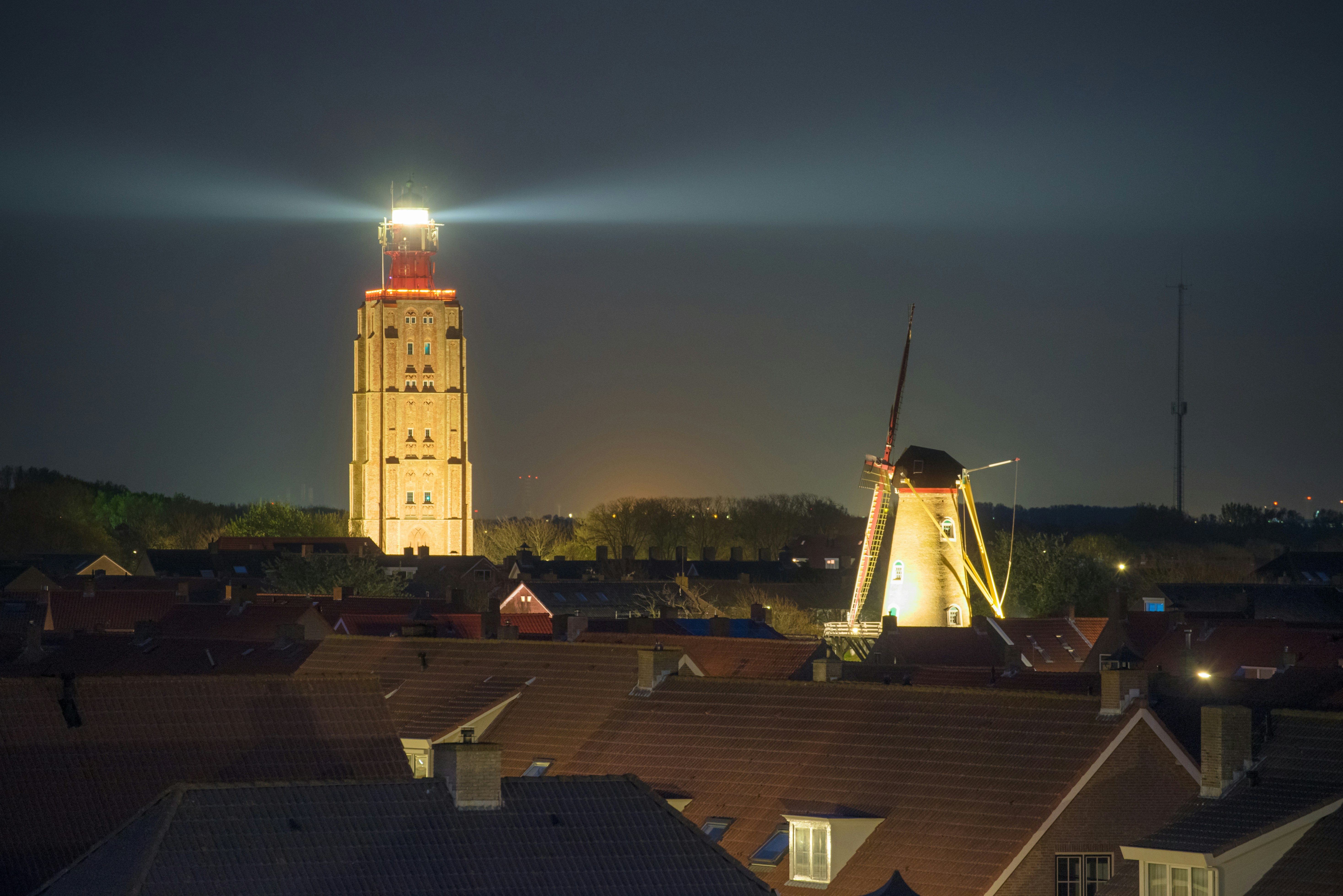 A lighthouse and windmill illuminated against the night sky, showcasing their architectural beauty amidst a tranquil urban landscape.