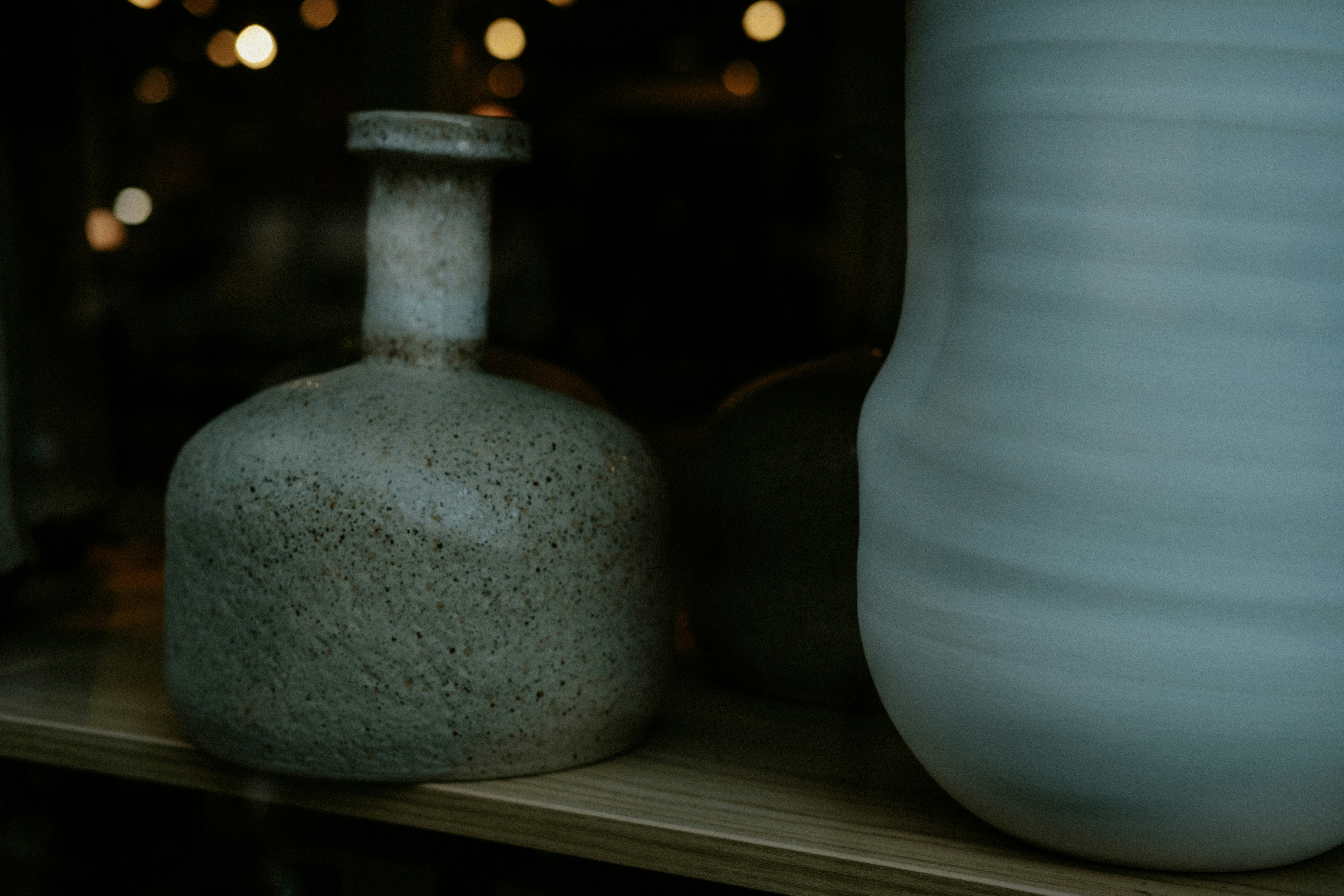 Two ceramic vessels, one textured and round, the other smooth and tall, displayed on a wooden shelf with soft, ambient lighting.