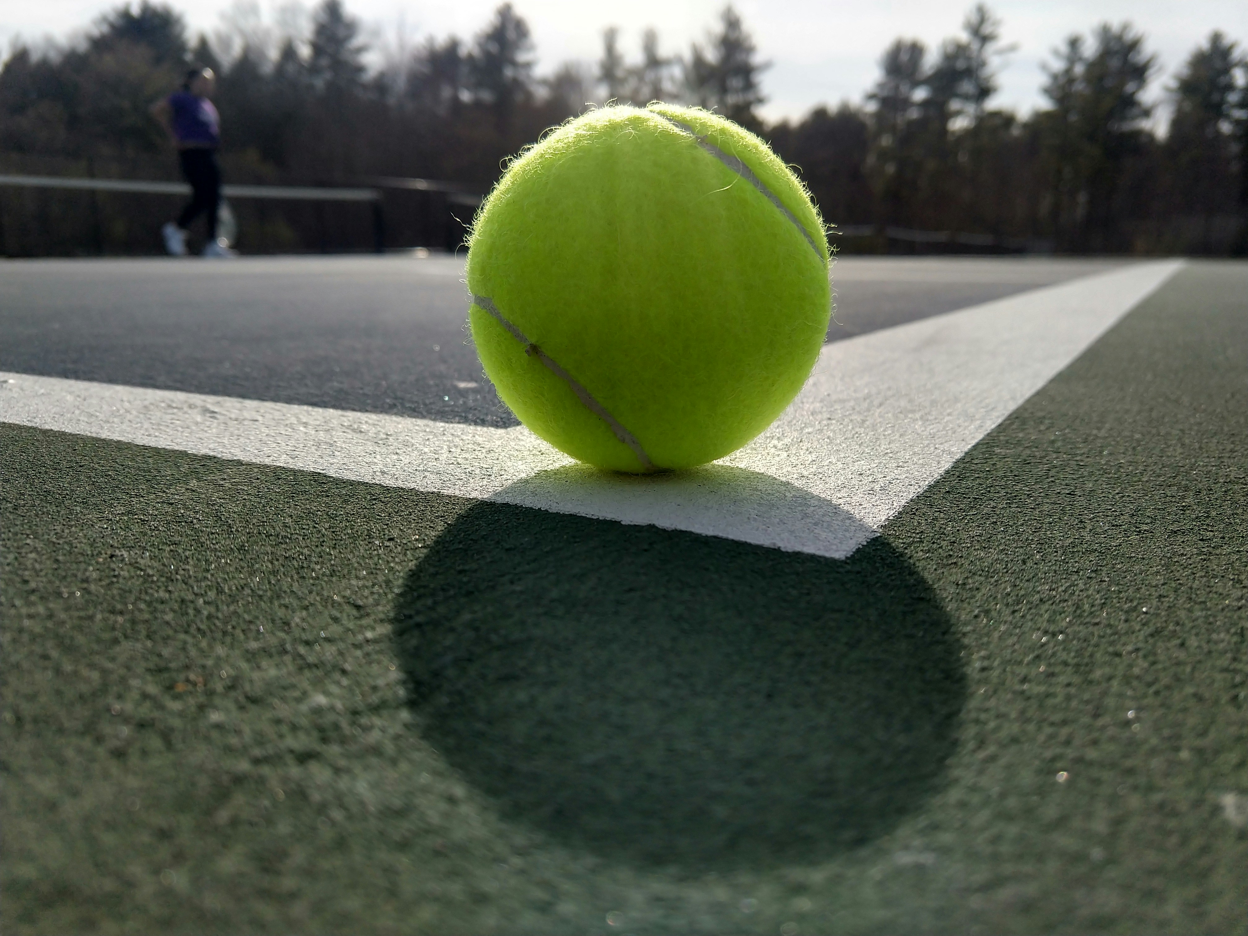 Vibrant green tennis ball resting on the court, casting a shadow on the painted lines, with a player in the background preparing for a match.