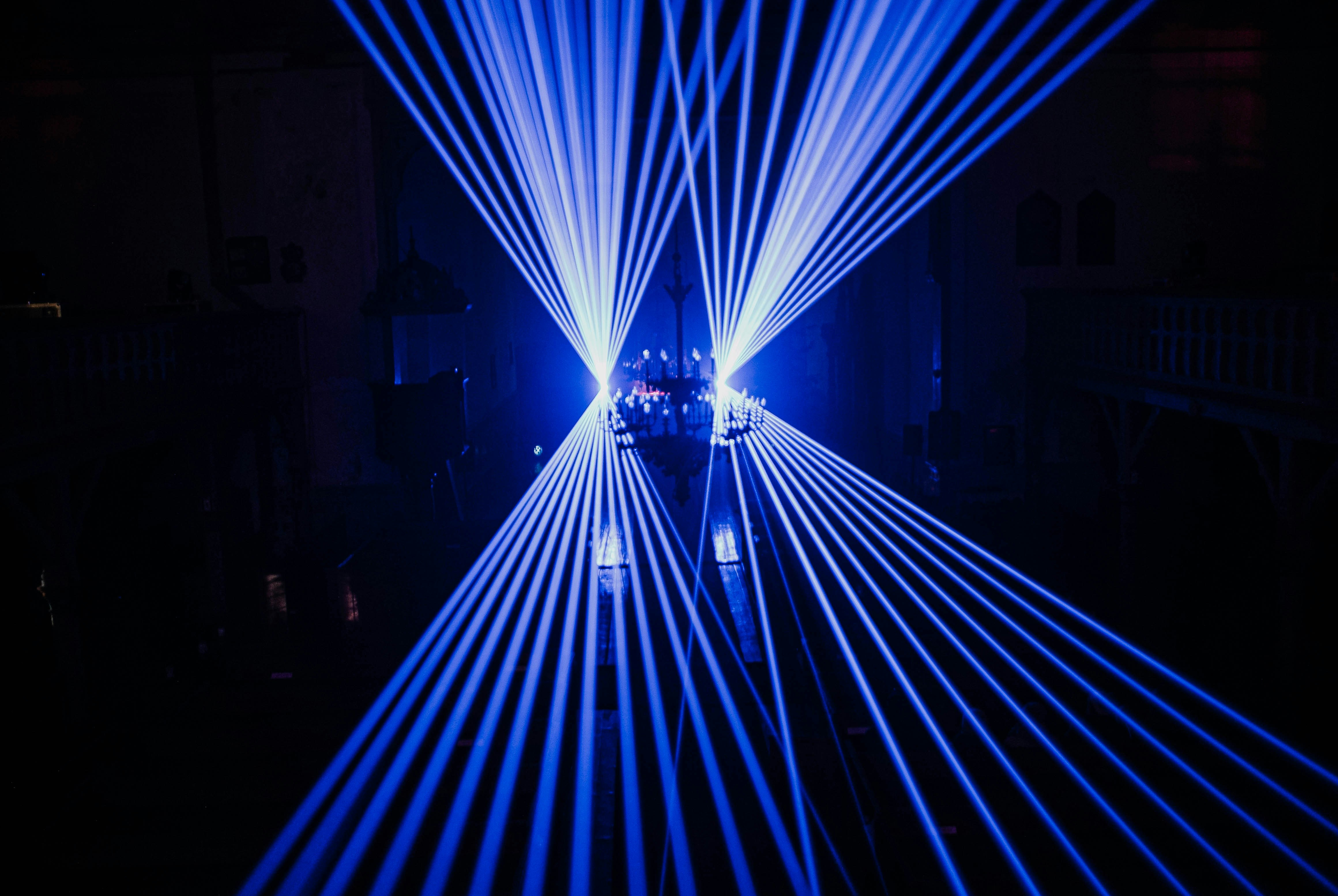 Vibrant blue laser beams converge dramatically in a darkened space, creating a mesmerizing visual spectacle. The scene captures the energy of a live performance.
