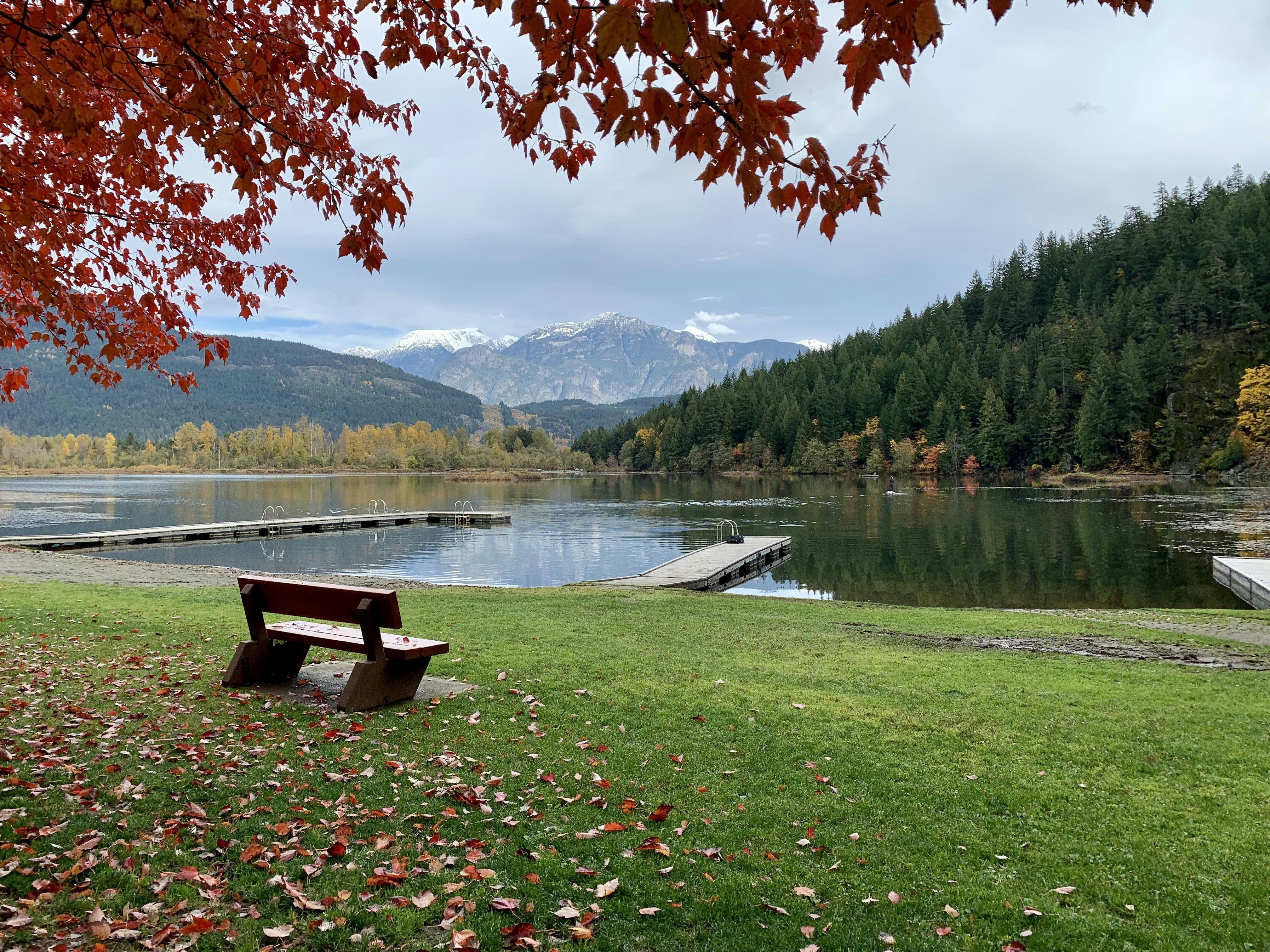 A tranquil lakeside scene framed by vibrant autumn leaves, with wooden docks extending into the water and mountains in the background.