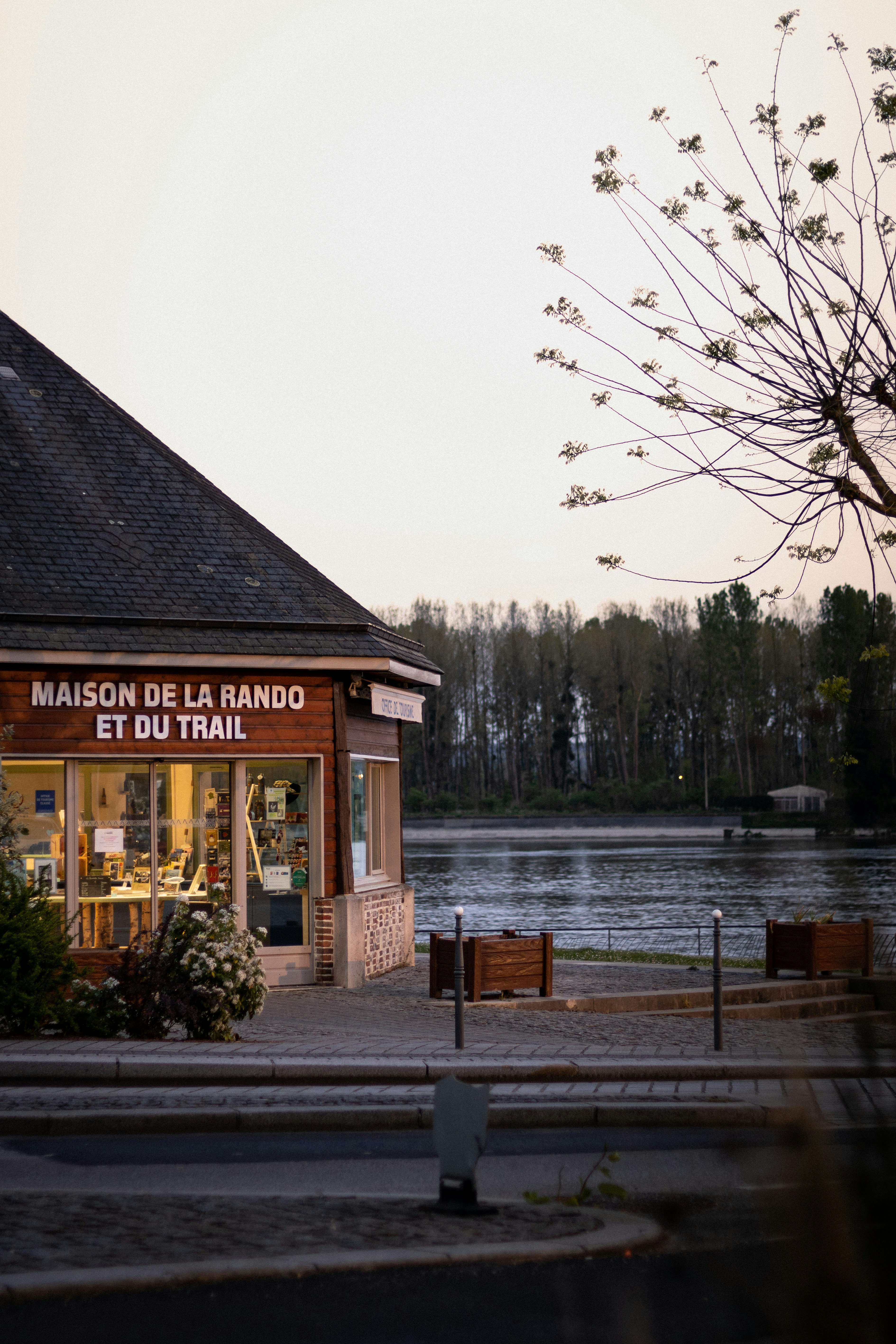Wooden building labeled 'MAISON DE LA RANDO ET DU TRAIL' beside a tranquil river, surrounded by trees and benches.
