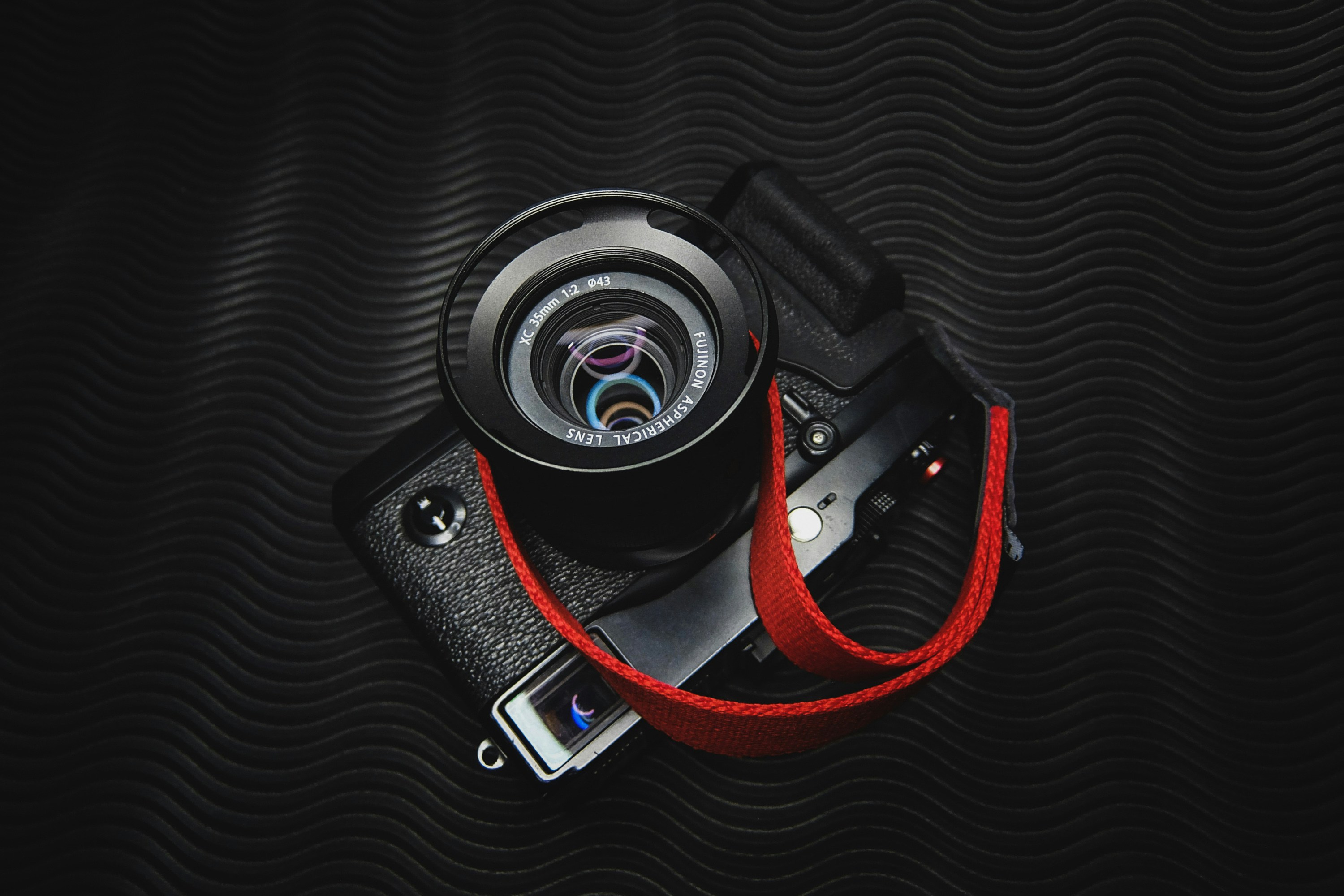 A compact digital camera with a prominent lens and red strap rests on a dark, wavy-textured backdrop.