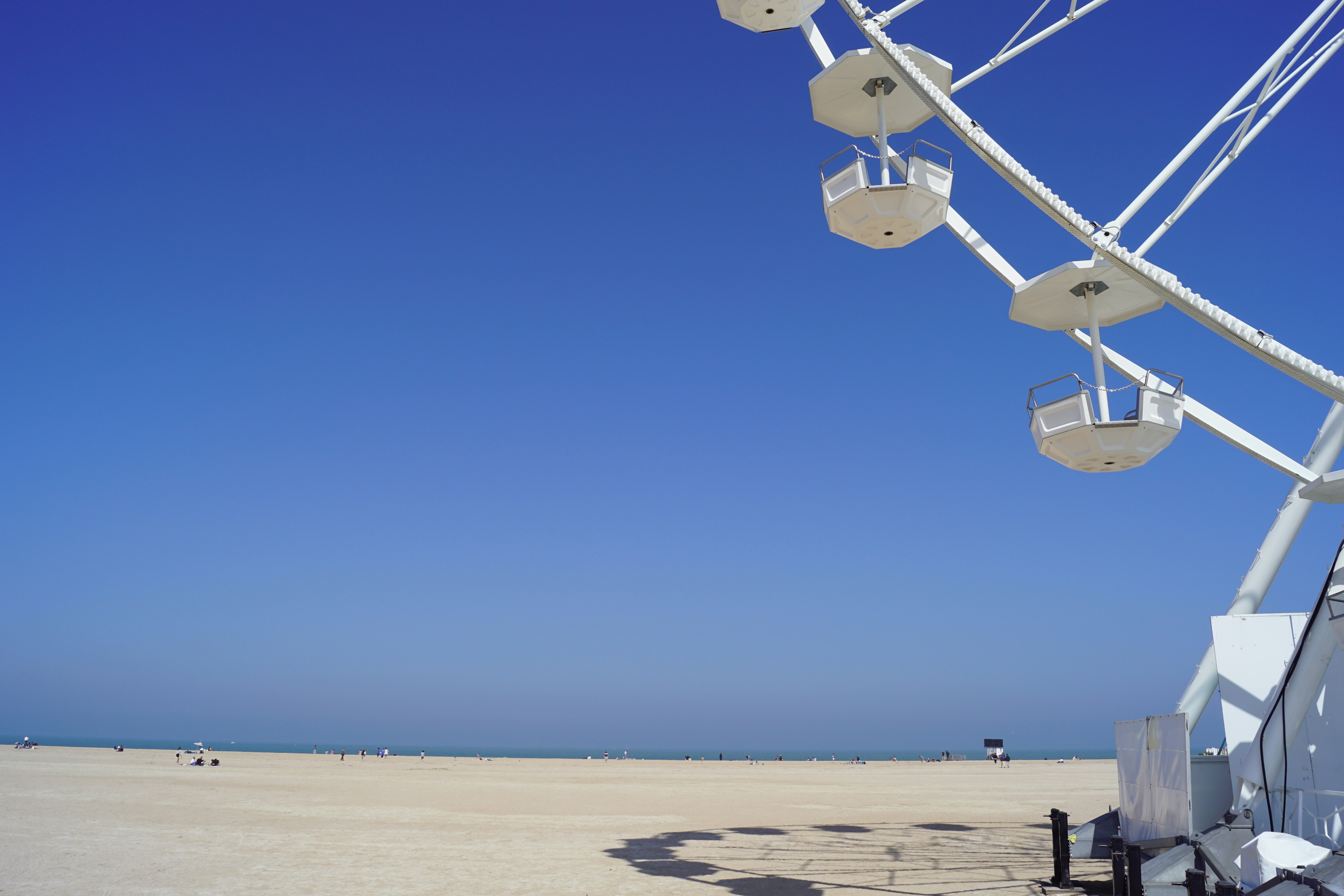 A towering Ferris wheel stands against a clear blue sky, overlooking an expansive beach scene with distant figures enjoying the sun.