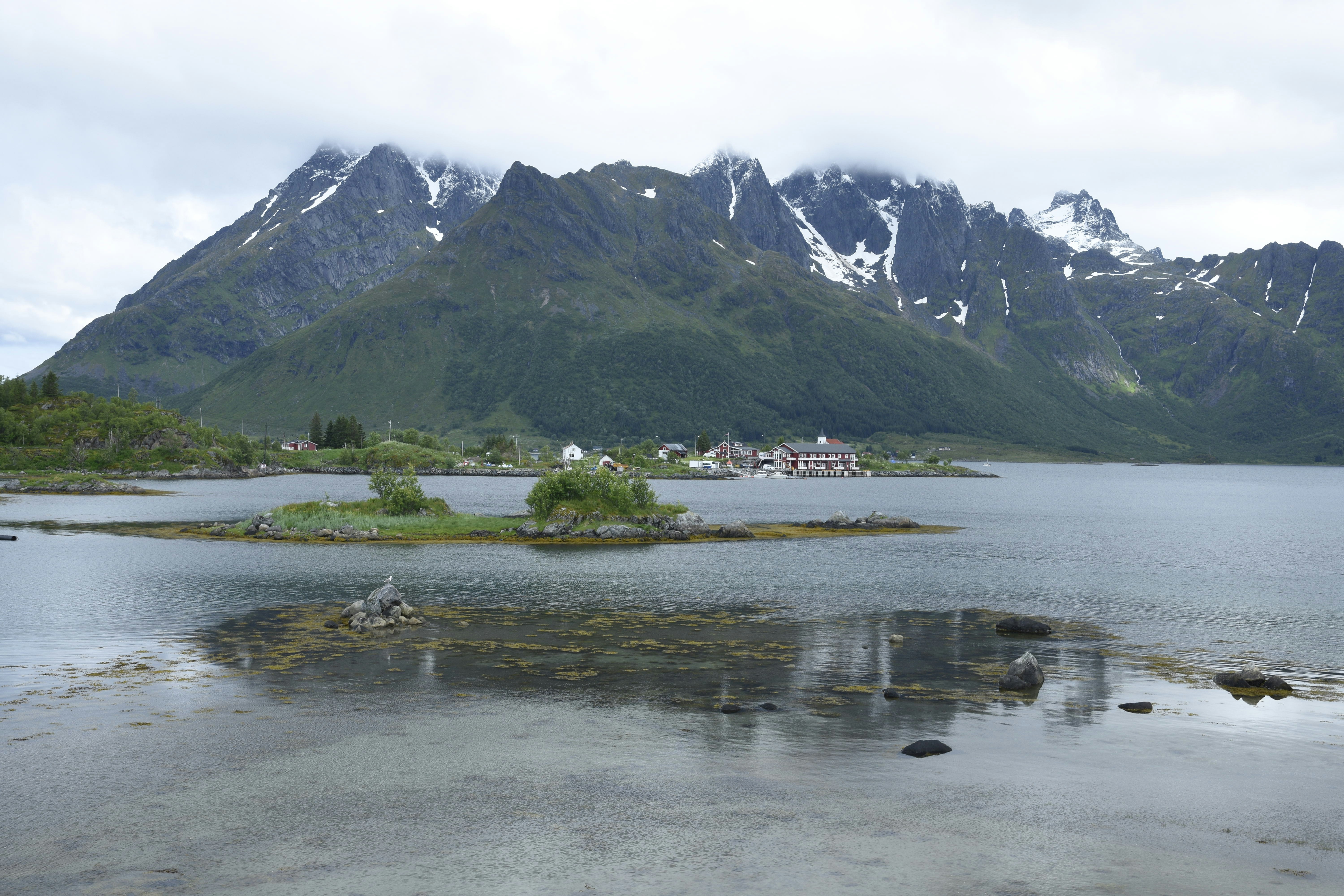 A tranquil coastal village nestled against towering mountains, reflecting on calm waters under a cloudy sky.