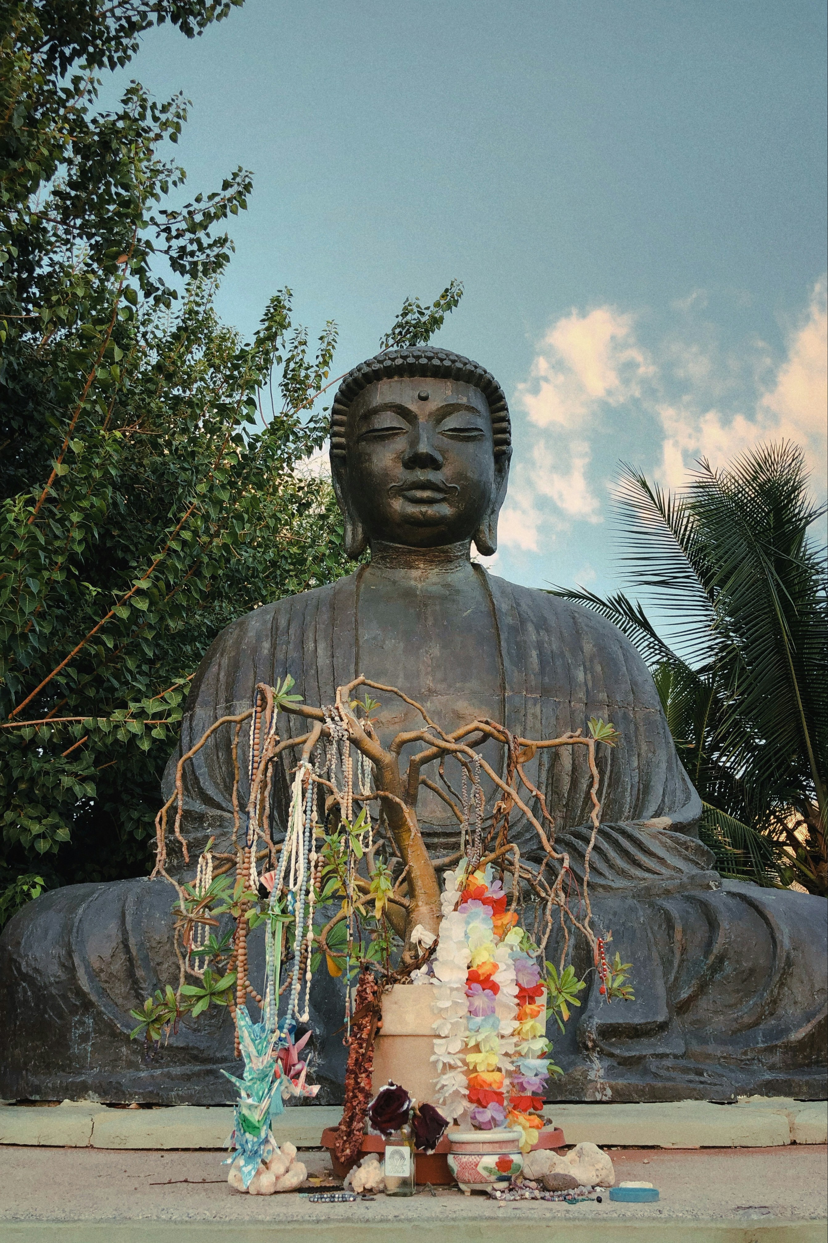 Majestic Buddha statue adorned with colorful offerings, surrounded by lush greenery and a serene sky. The scene evokes tranquility and reverence.