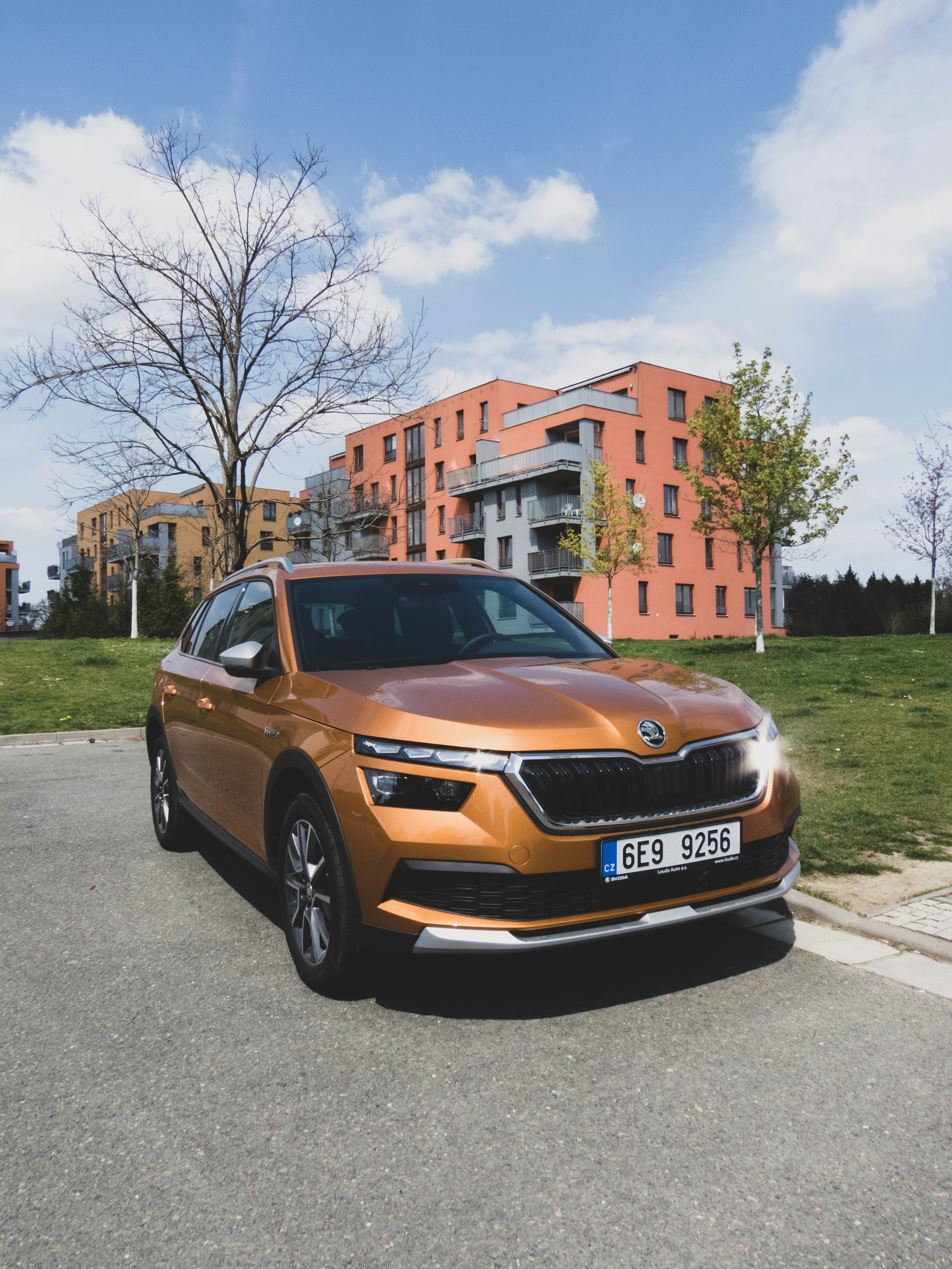 Orange compact SUV parked on a quiet street with contemporary buildings in the background. The scene captures a blend of urban architecture and automotive design.
