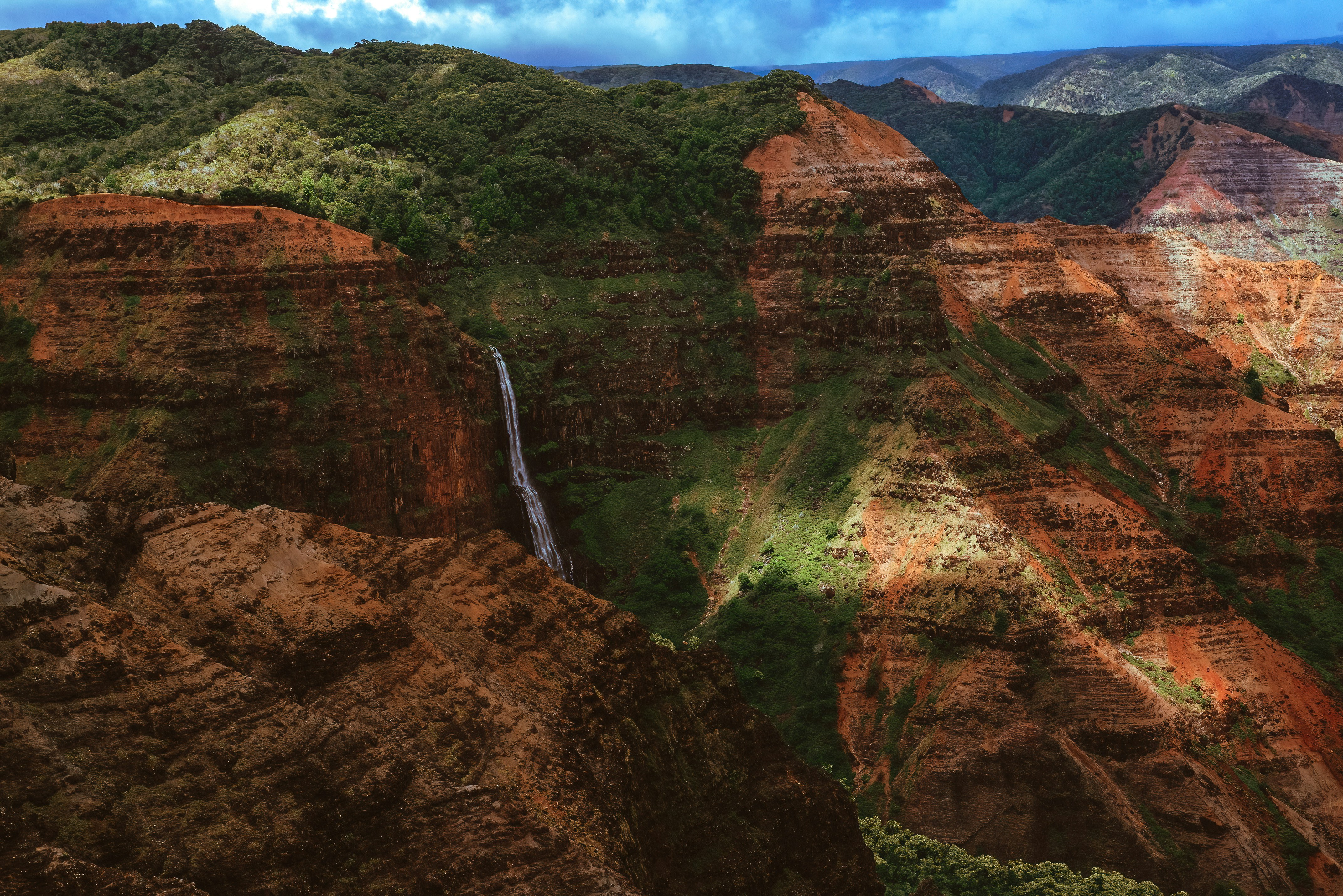 A cascading waterfall flows down vibrant red cliffs, surrounded by lush greenery in a dramatic canyon landscape.