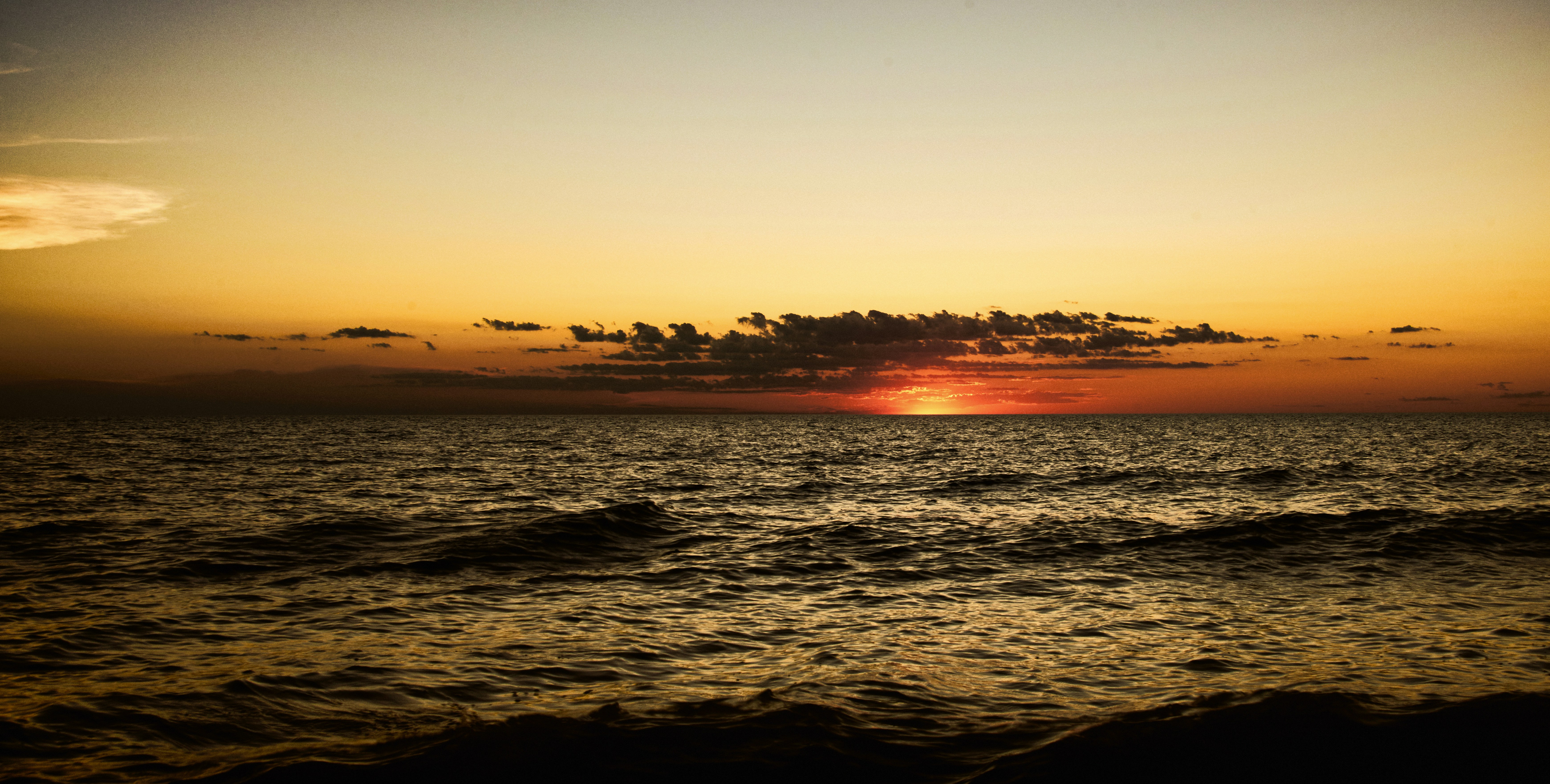 Golden sun dipping below the horizon, casting warm reflections on the gentle ocean waves. The serene atmosphere captures the transition from day to night.