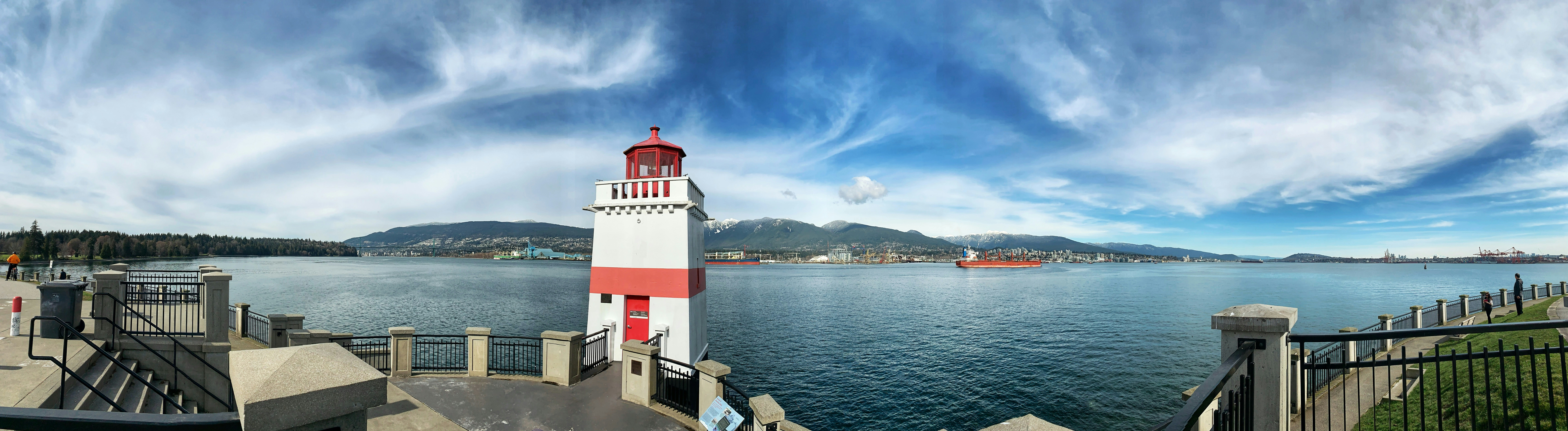Panoramic view of a red and white lighthouse by the water with distant mountains under a dramatic sky.