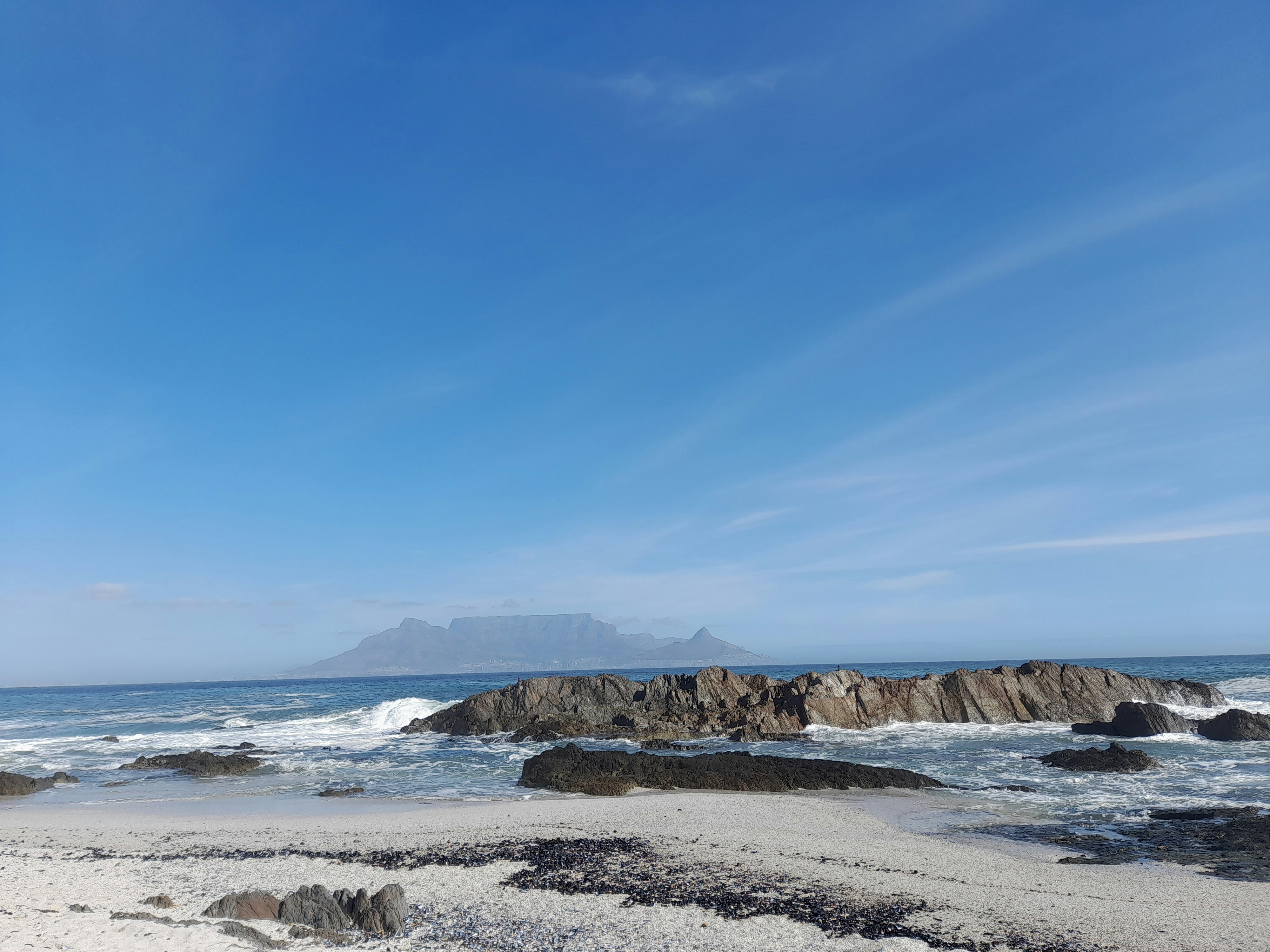 Rocky shoreline with waves breaking over jagged rocks. A distant flat-topped landmass sits on the horizon beneath a crisp blue sky.