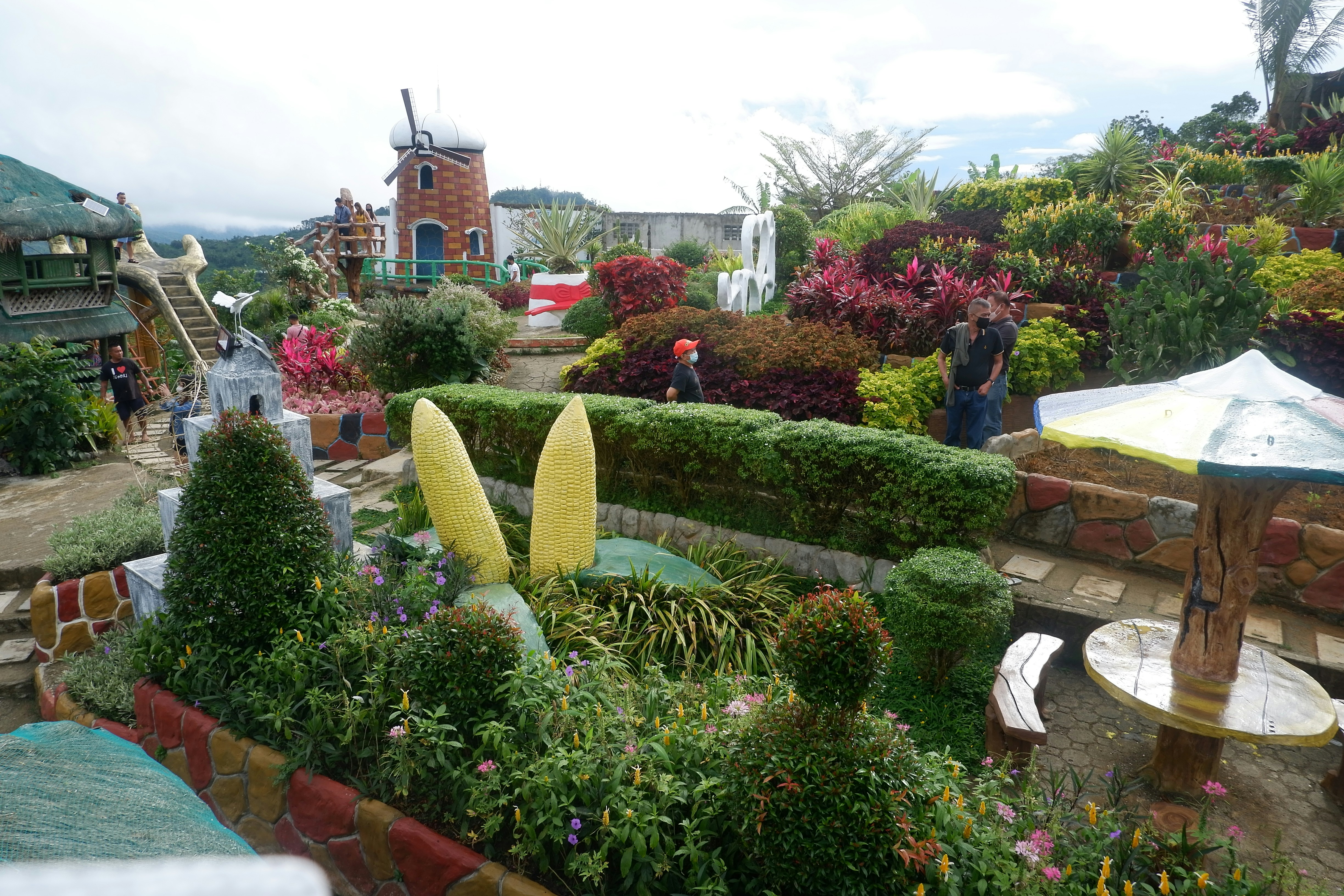 A colorful, neatly manicured garden labyrinth with winding hedges, sculpted topiary, and playful architectural features beneath a bright sky.