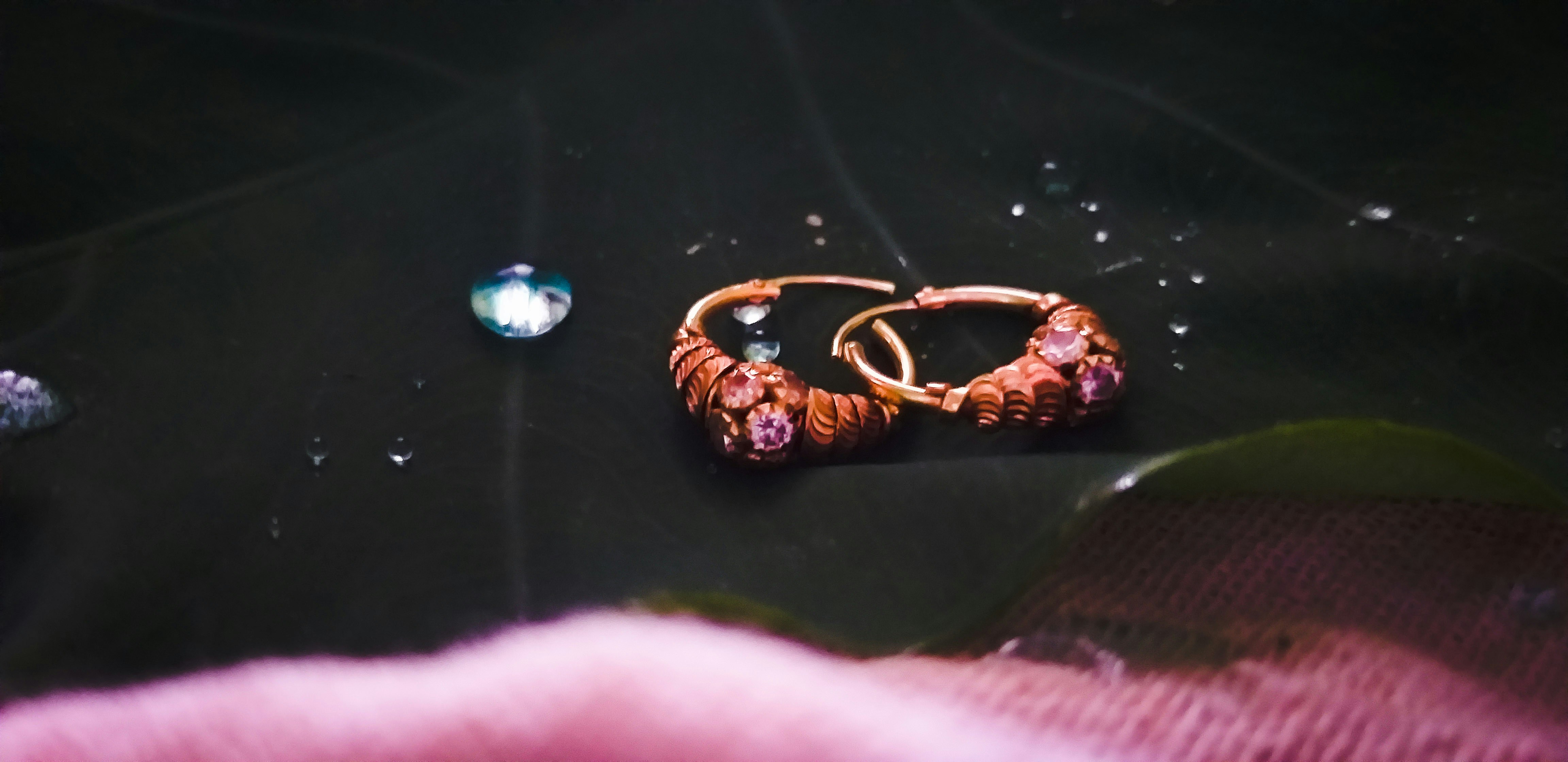 Delicate earrings resting on a dark surface, surrounded by droplets of water and soft fabric. The intricate design shines subtly in the light.
