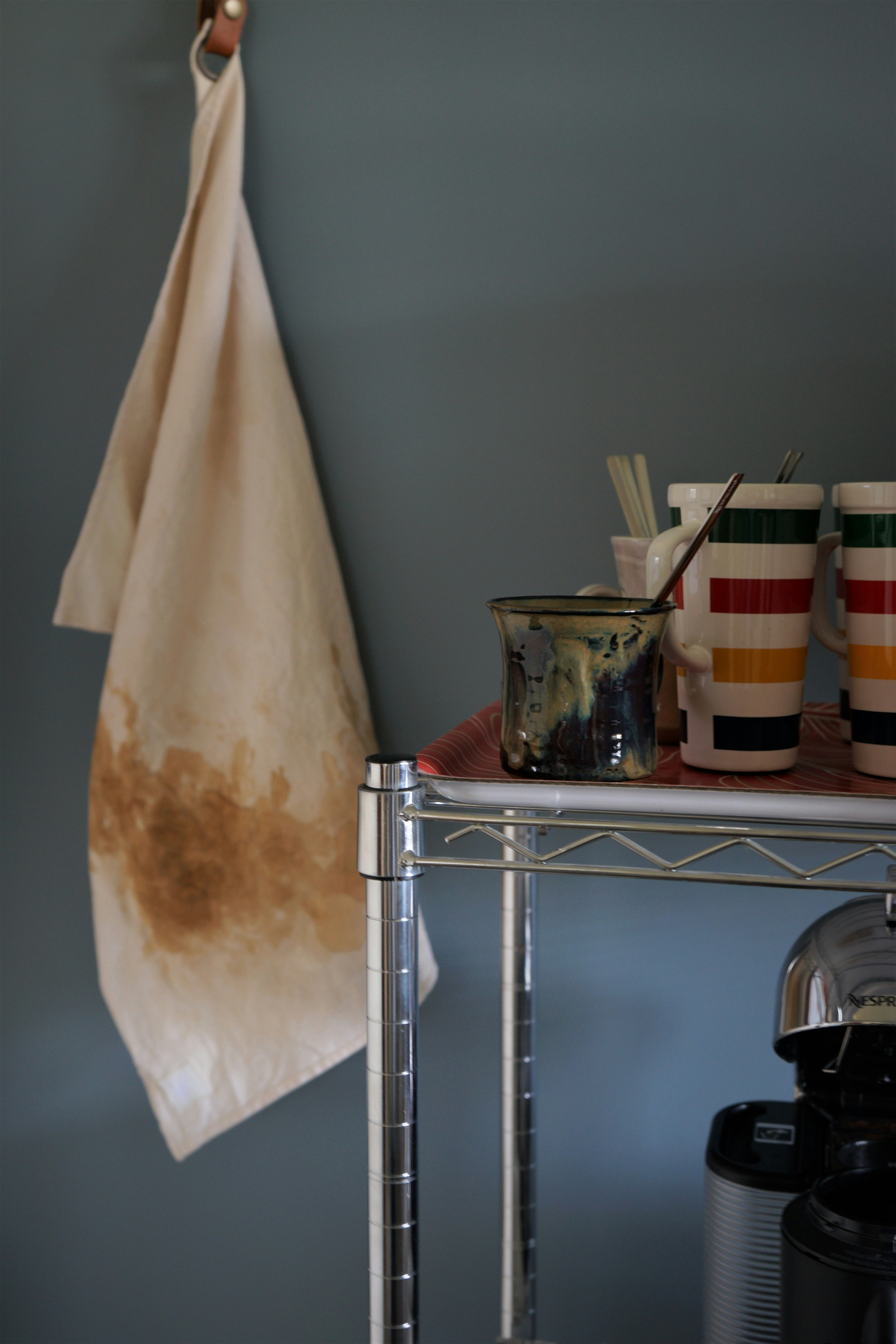 Muted kitchen vignette: stained towel beside striped mugs on chrome shelf