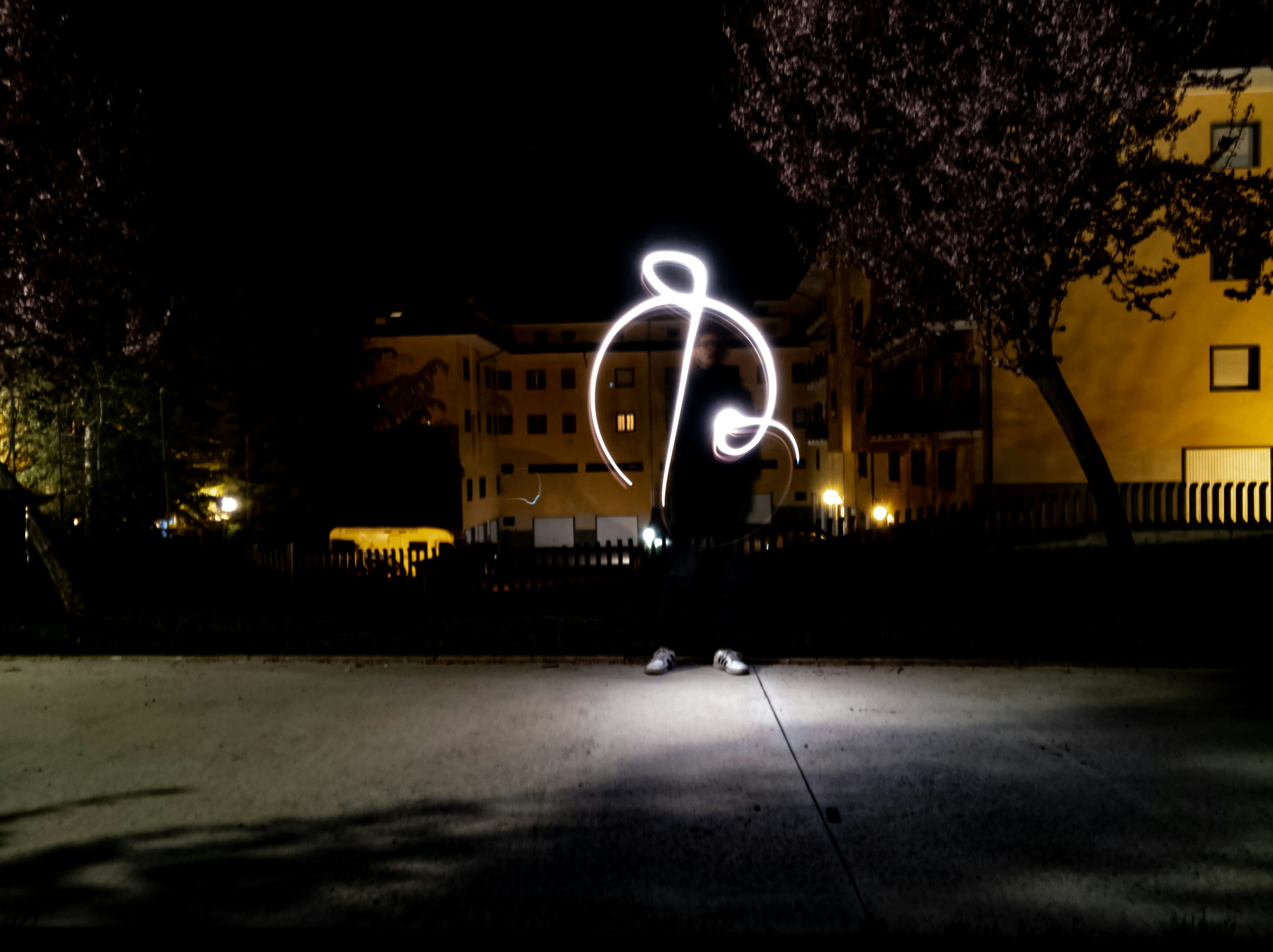 Long-exposure night photograph capturing a person weaving a neon light trail in a quiet park. Soft ground illumination and distant building silhouettes frame the luminous arc around the mover.