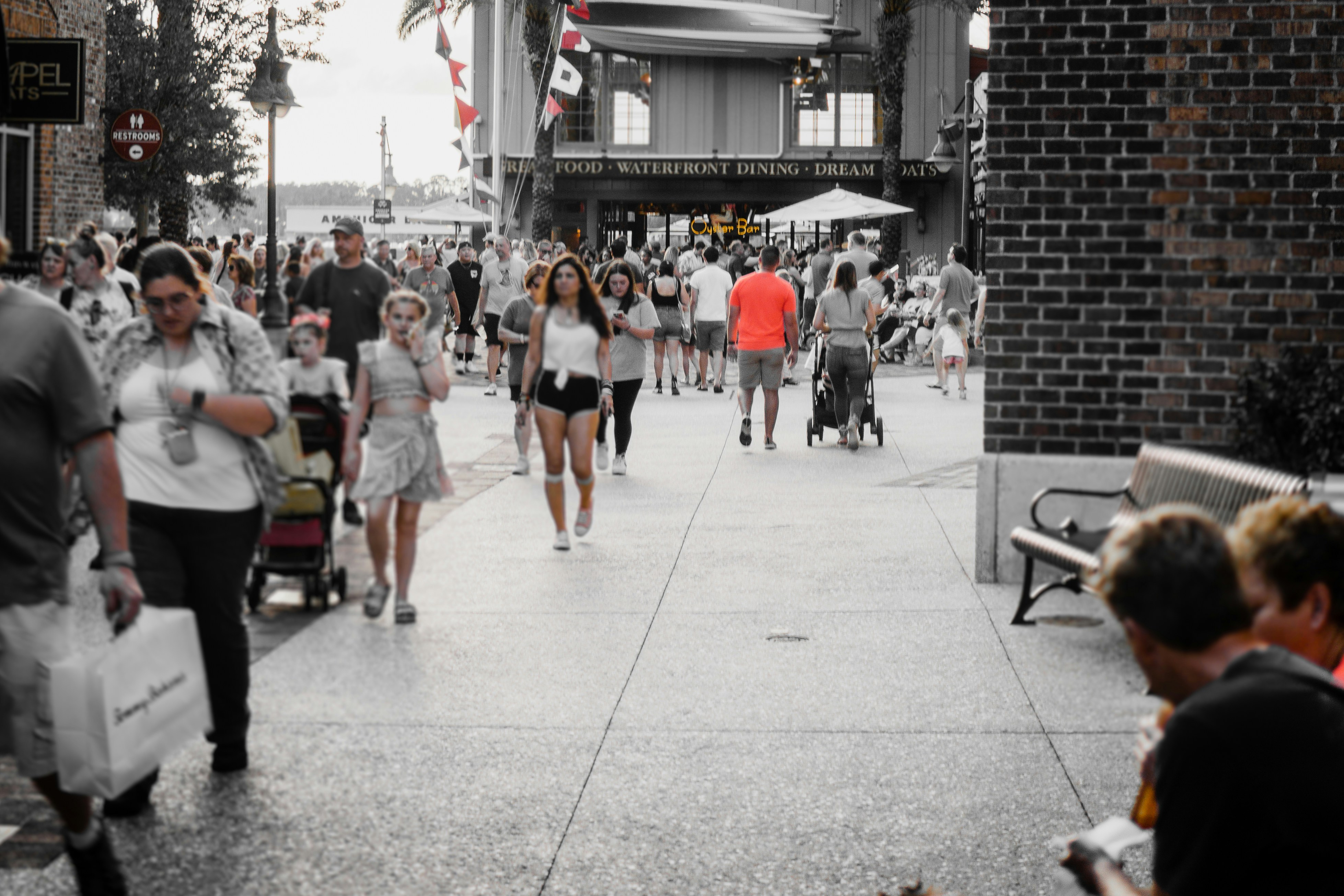 Busy waterfront promenade filled with pedestrians, featuring a striking contrast between colorful and monochrome elements. A family strolls alongside others enjoying the lively atmosphere.