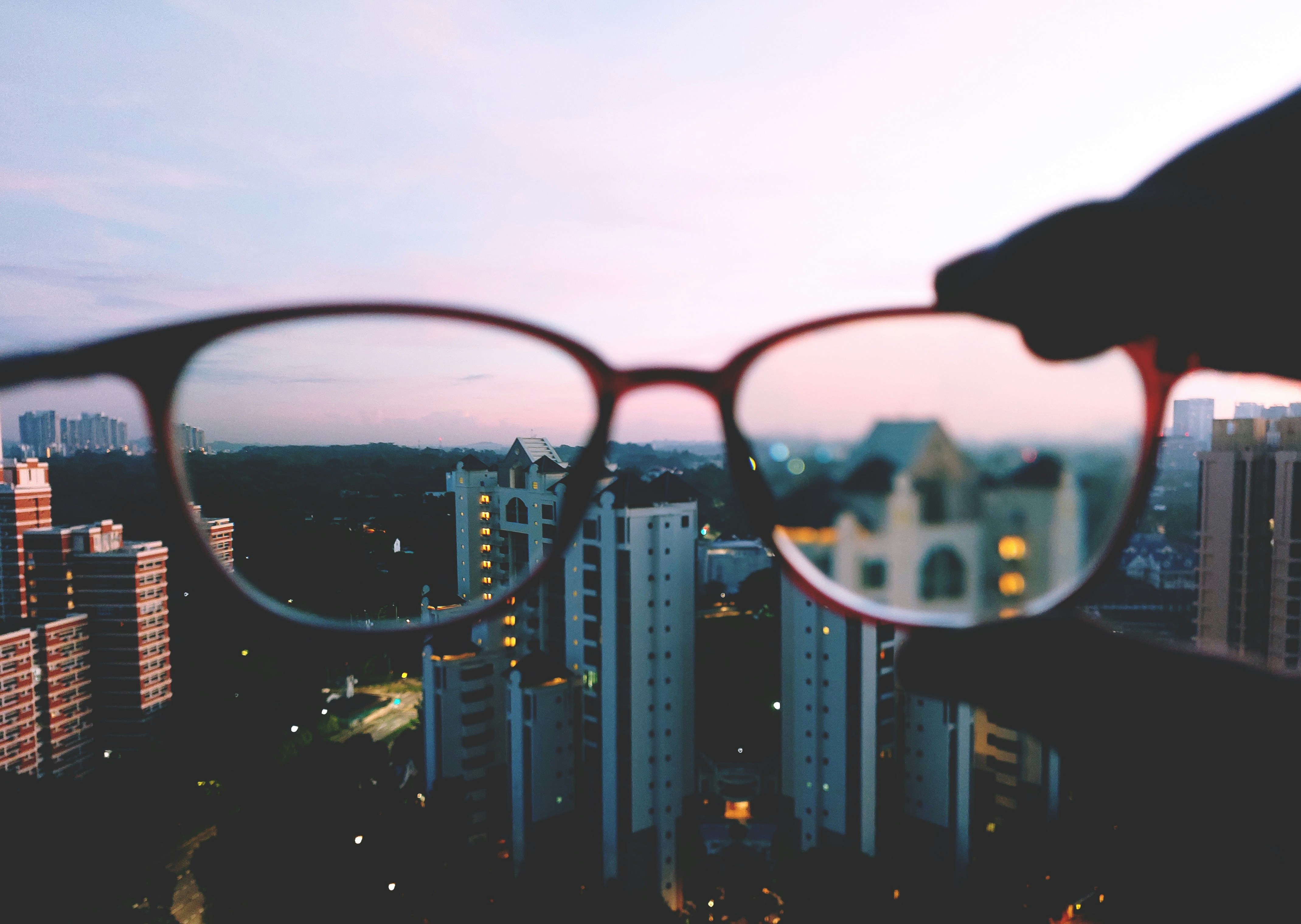 A pair of glasses held in front of a cityscape, highlighting the contrast between the blurred foreground and the detailed buildings in the background.